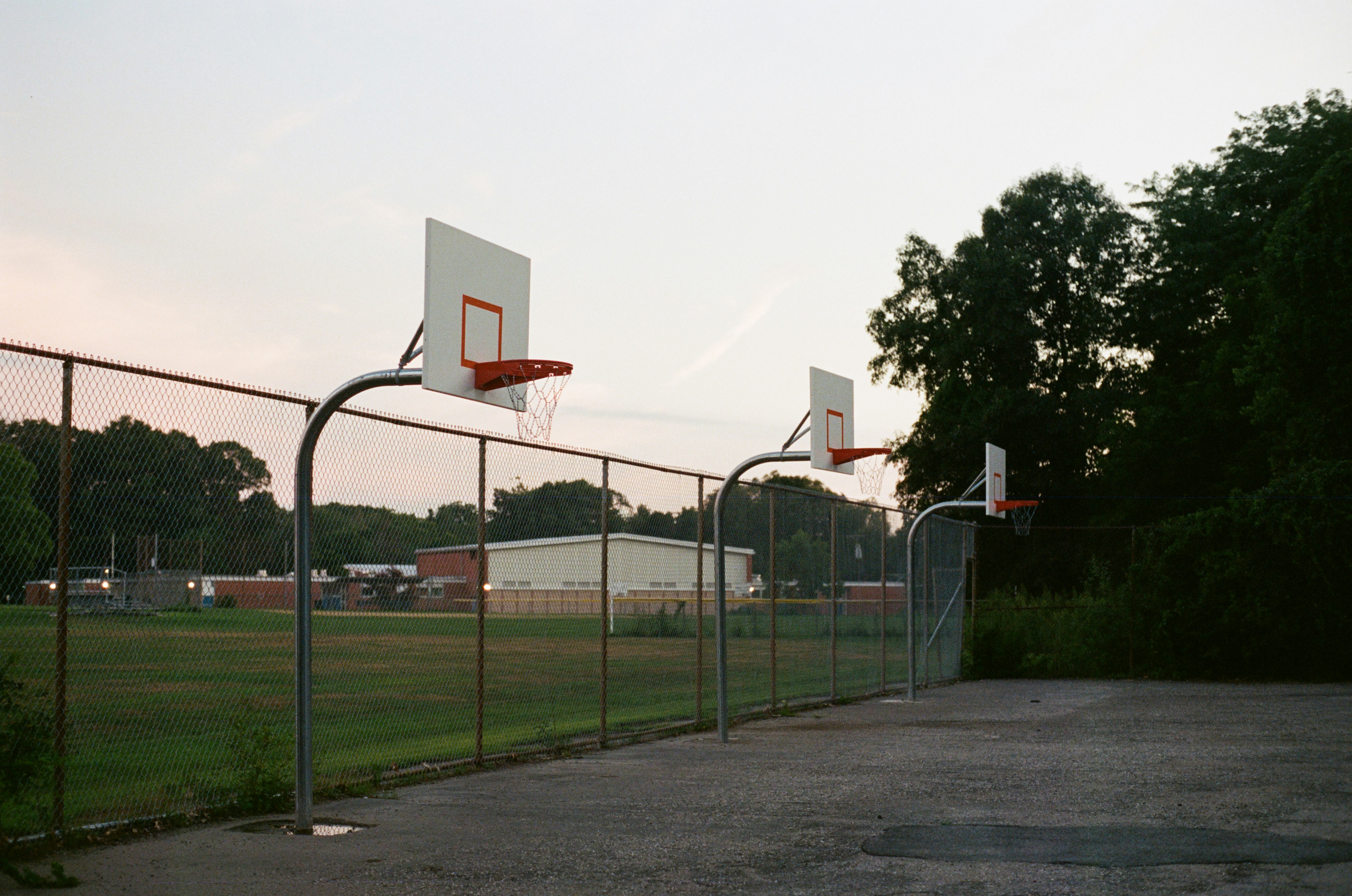 Abandoned basketball court with empty hoops silhouetted against a fading sky, surrounded by a chain-link fence and lush greenery.