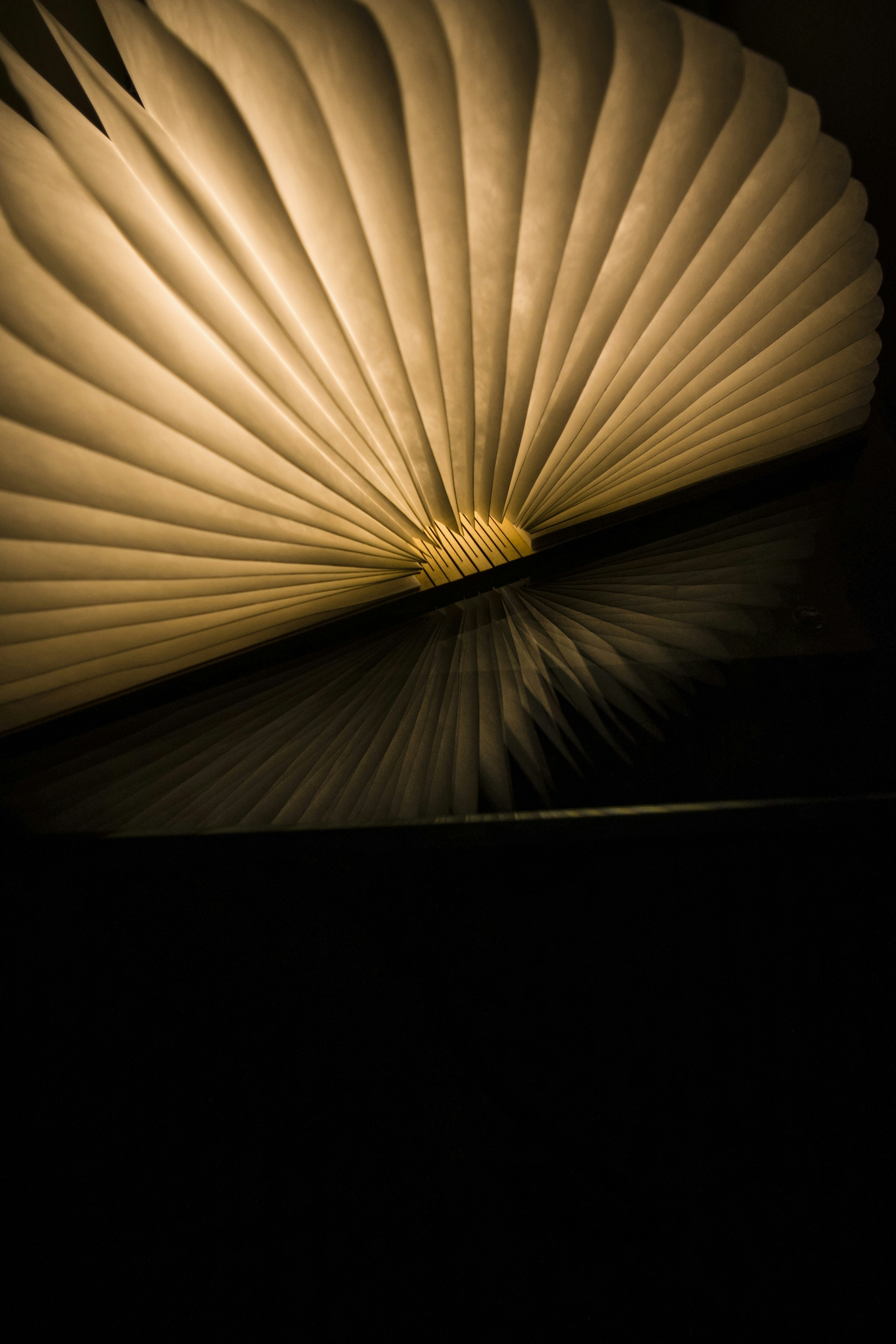 Close-up of a warm, pleated paper lamp shade lit from within, resting on a glossy surface. Its radial folds cast a soft amber glow and mirror-like reflection.