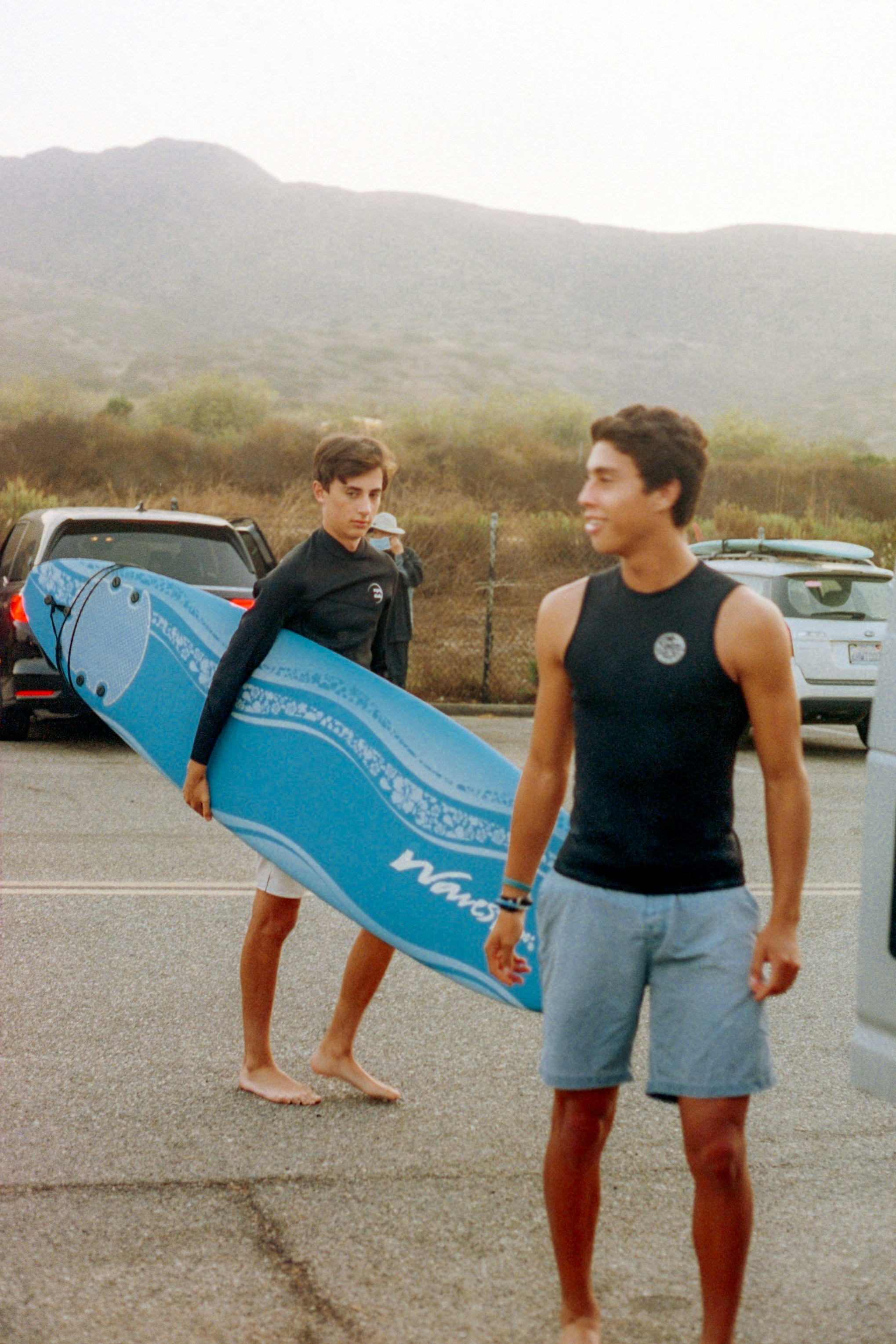 Two young surfers preparing for a day at the beach, one carrying a vibrant blue surfboard. The backdrop features rolling hills and parked cars.