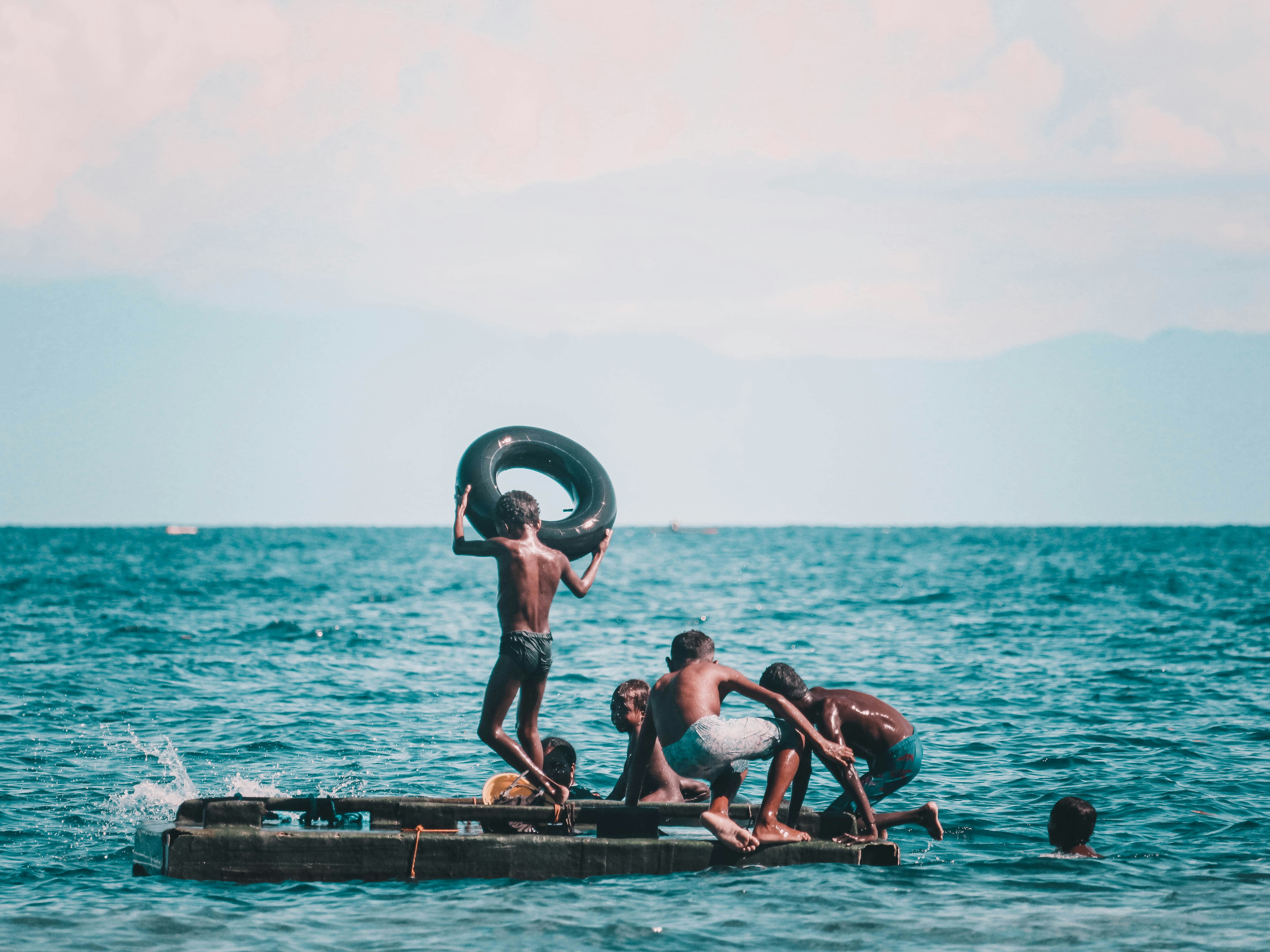 Children playing on a homemade raft in the ocean under a cloudy sky.