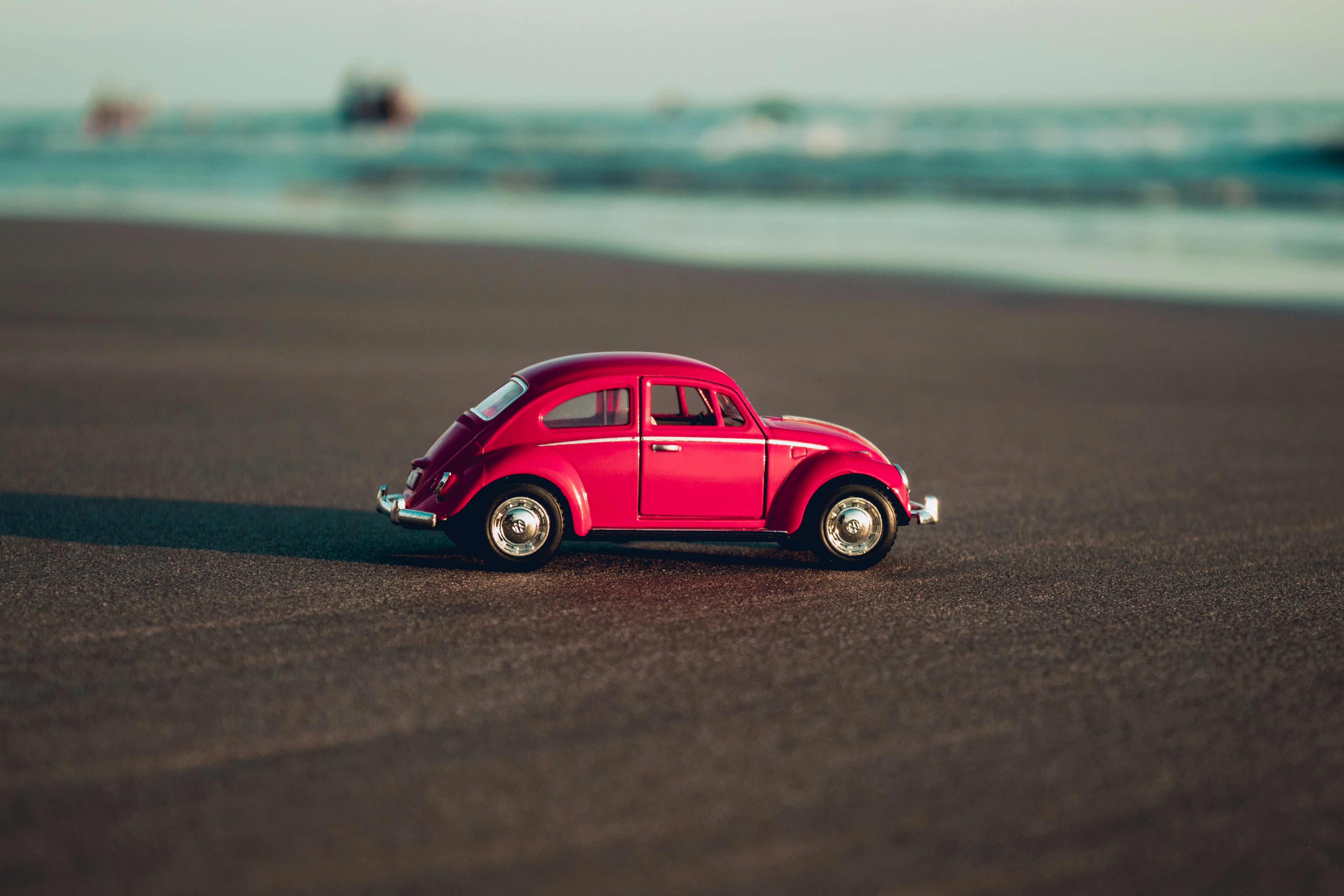 A vibrant red toy car positioned on a sandy beach, with gentle waves in the background. The scene captures a playful juxtaposition of nostalgia and nature.