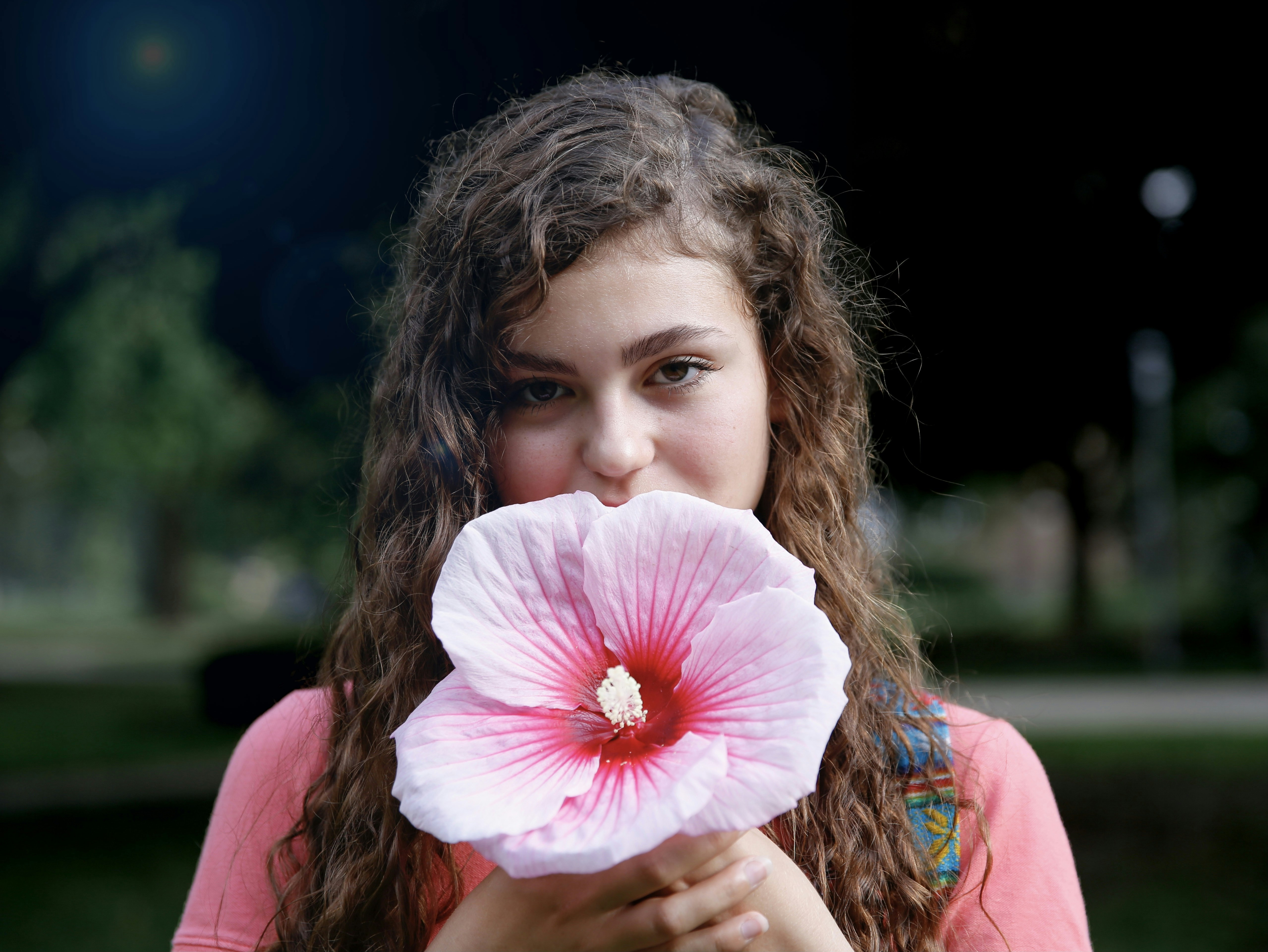 Girl in a pink shirt holding a large pink hibiscus flower close to her face.