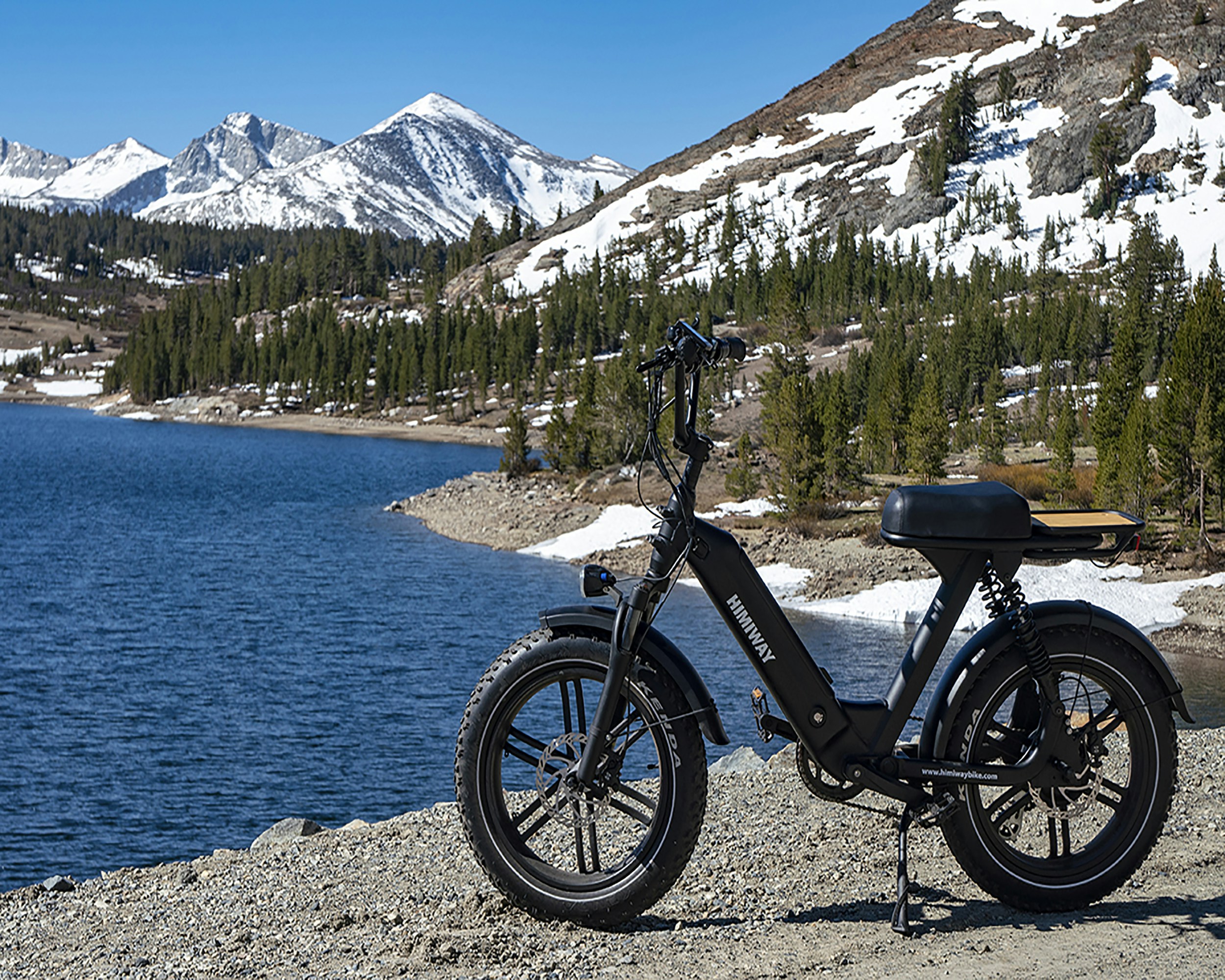 An electric bike parked by a tranquil lake, surrounded by snow-capped mountains and lush evergreen trees.
