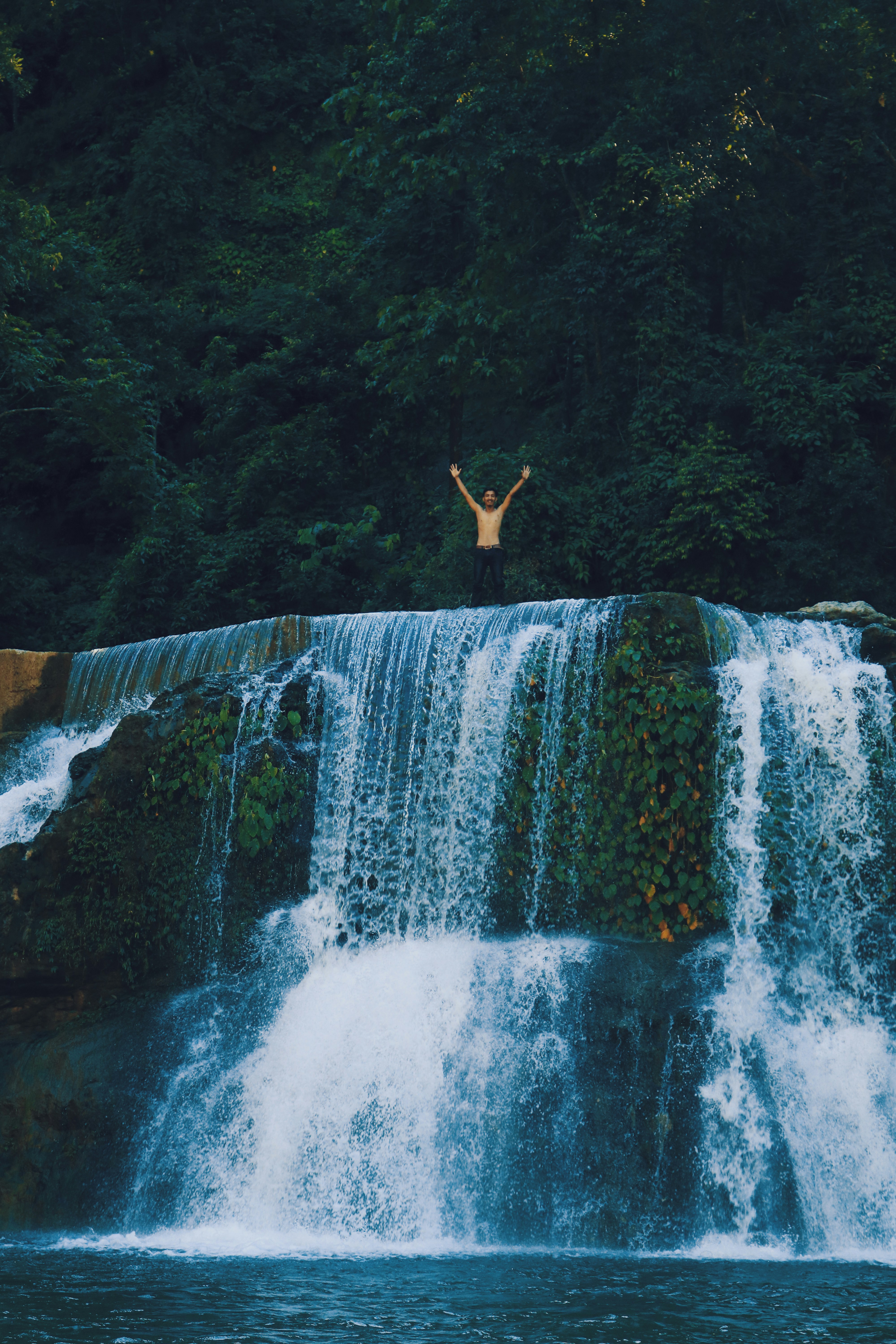 Person celebrating atop a waterfall surrounded by lush greenery. The cascading water creates a vibrant backdrop.