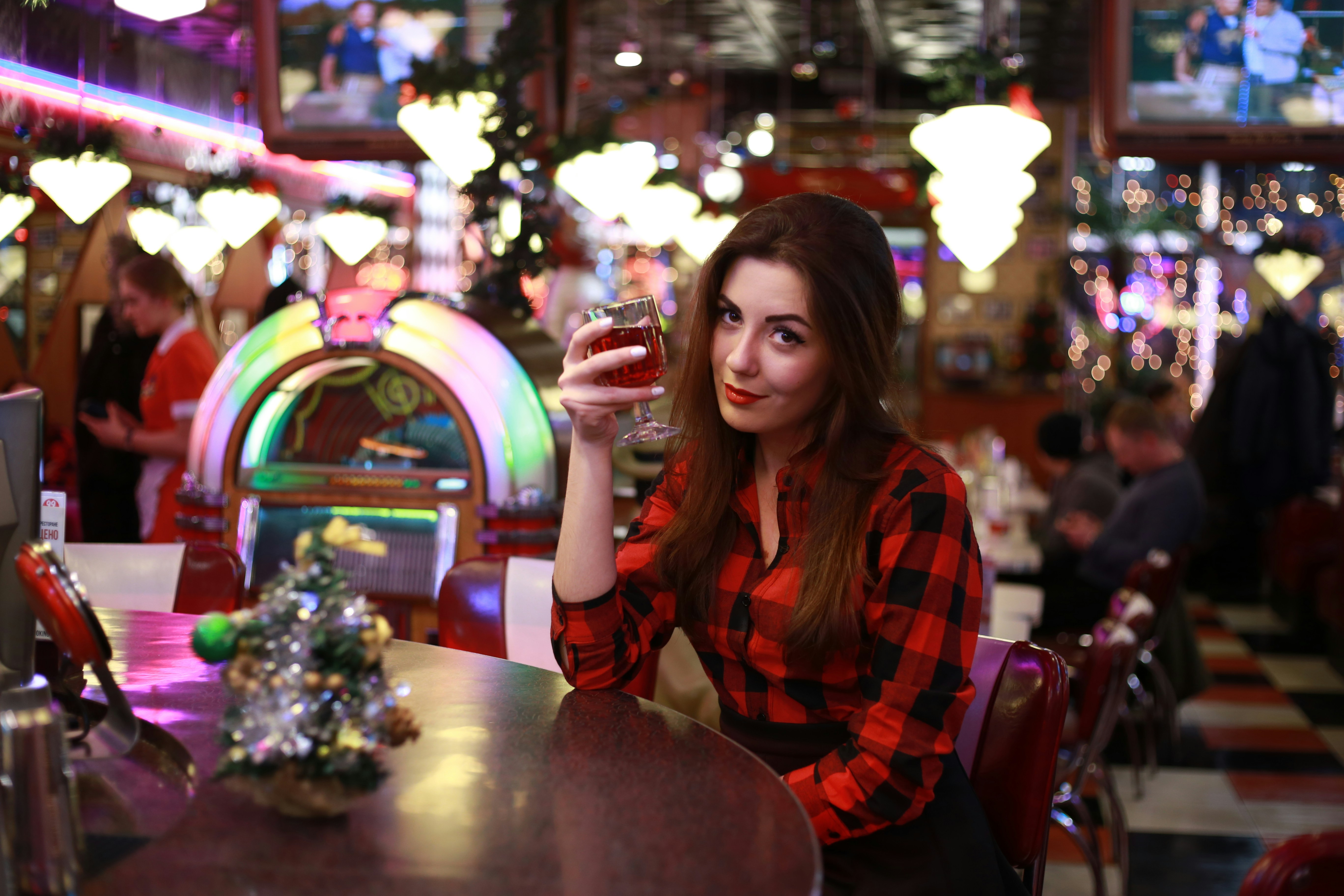 Woman in a plaid shirt raises a glass at a retro diner, surrounded by colorful decor and vintage elements. The scene captures a blend of modern and nostalgic ambiance.