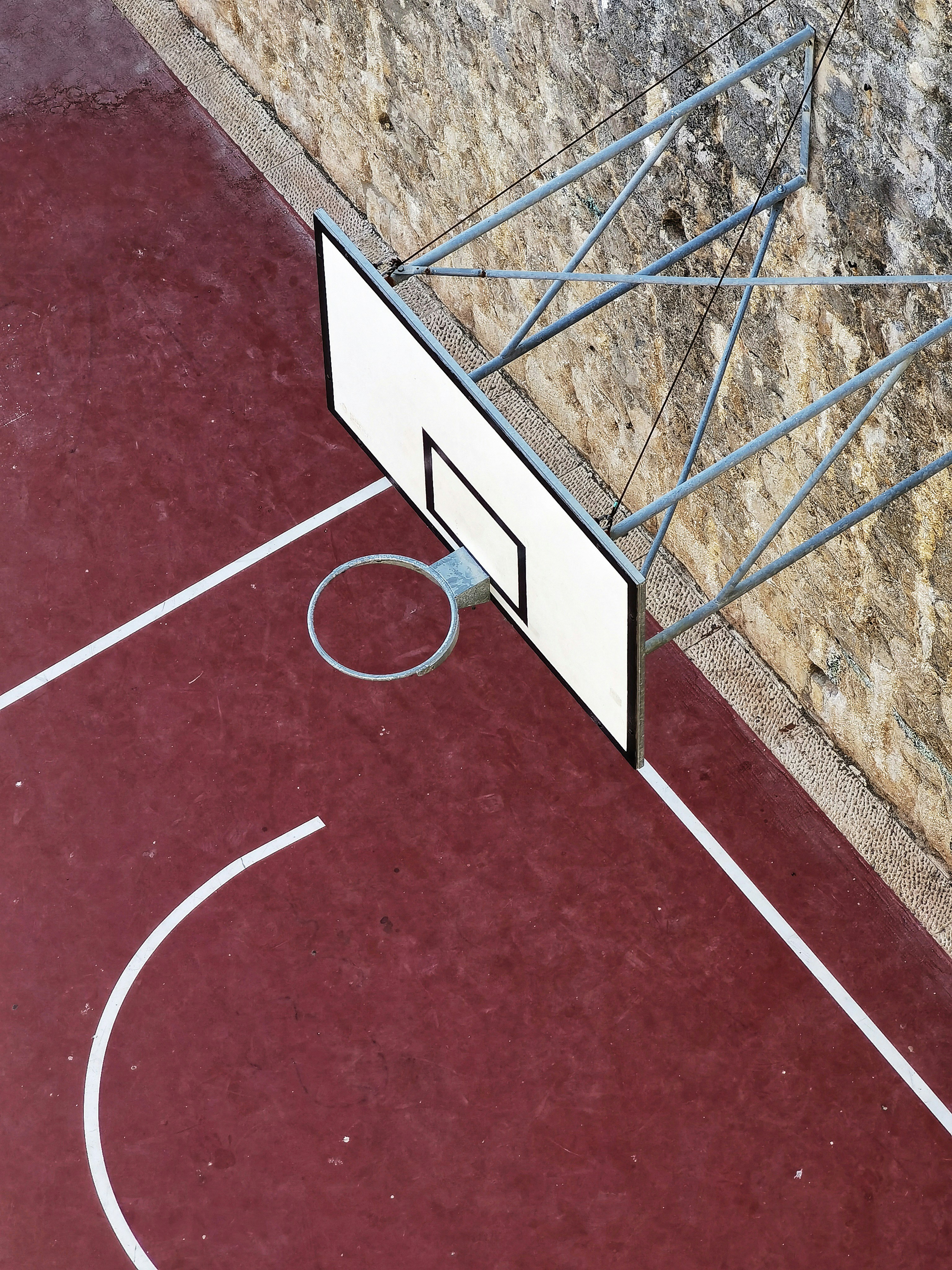 Basketball hoop mounted on a weathered stone wall, with a red court surface and white markings. The perspective highlights the geometric patterns created by the court lines.