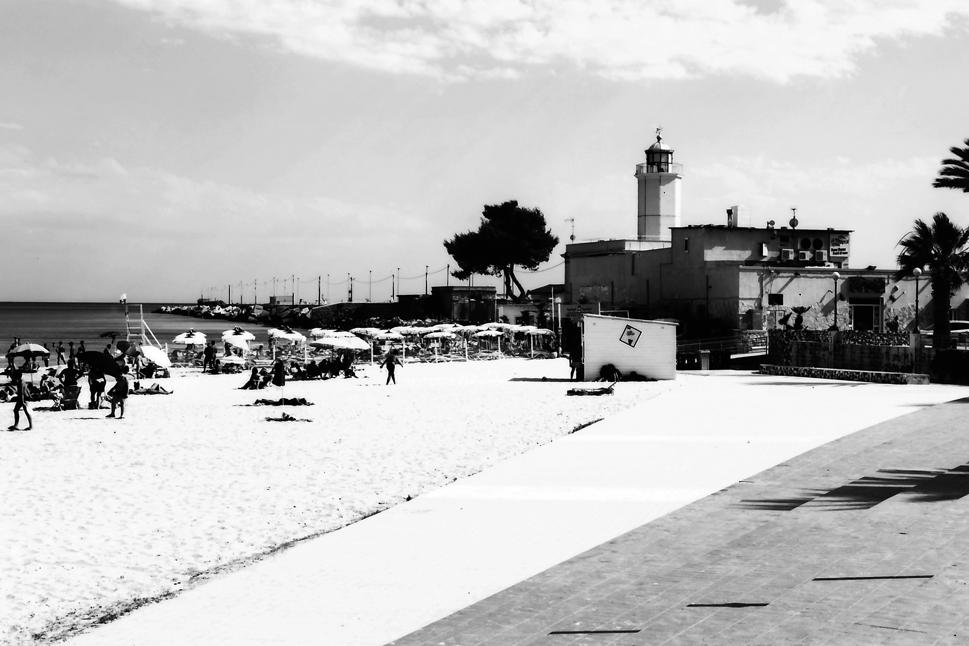 Lighthouse overlooking a bustling beach and port under a partly cloudy sky.