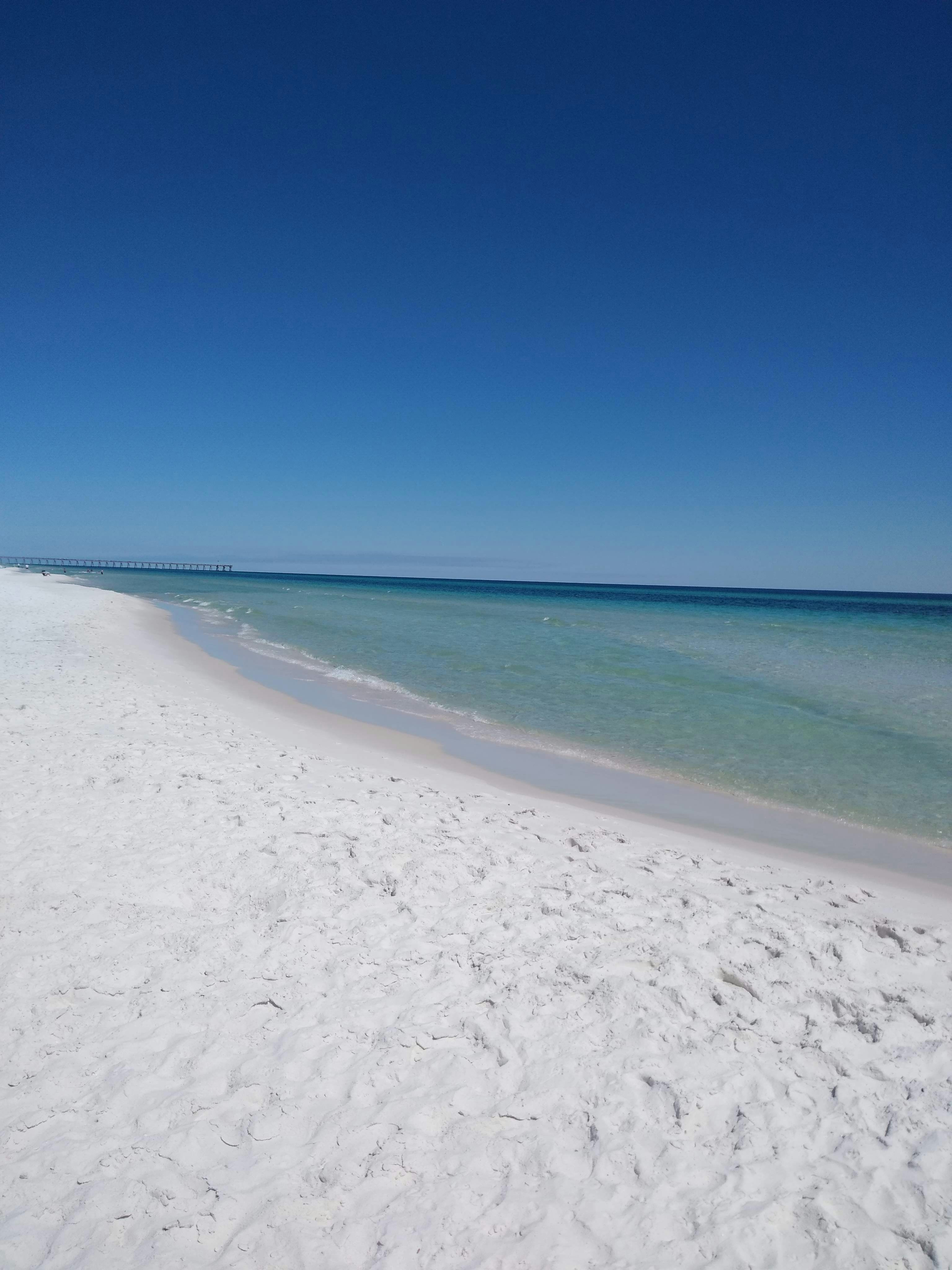 Tranquil beach scene featuring pristine white sand and clear turquoise waters under a bright blue sky. A distant pier adds depth to the horizon.