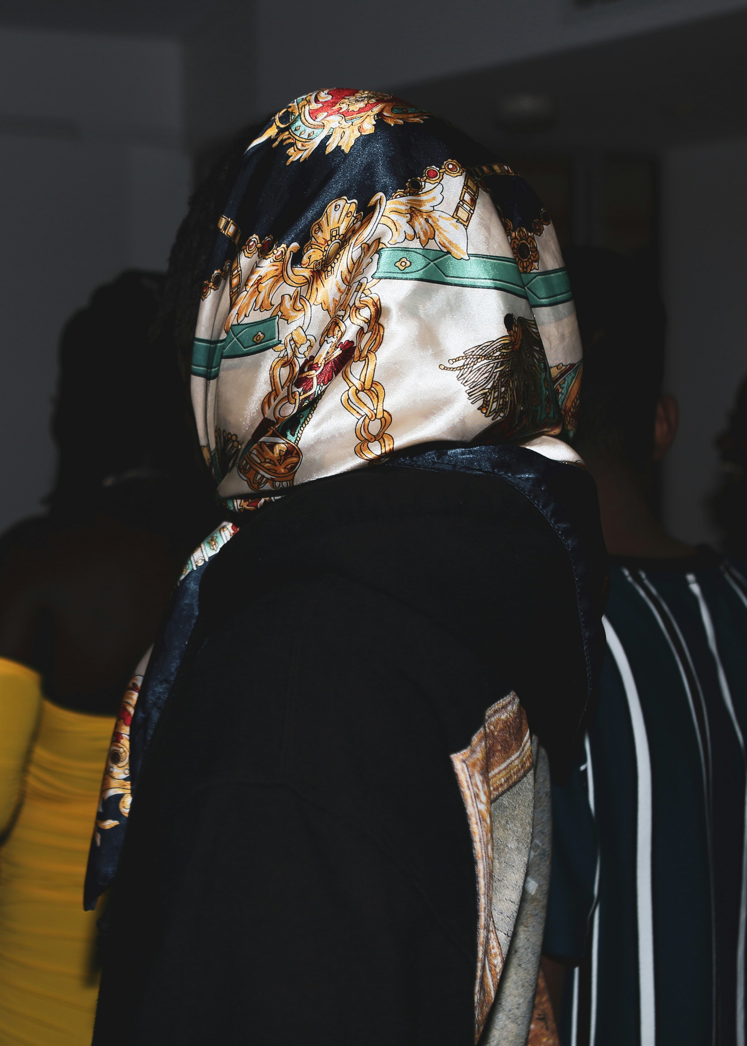 A figure adorned in a richly patterned scarf stands amidst a blurred background of colorful attire, capturing a moment of stylish anonymity.