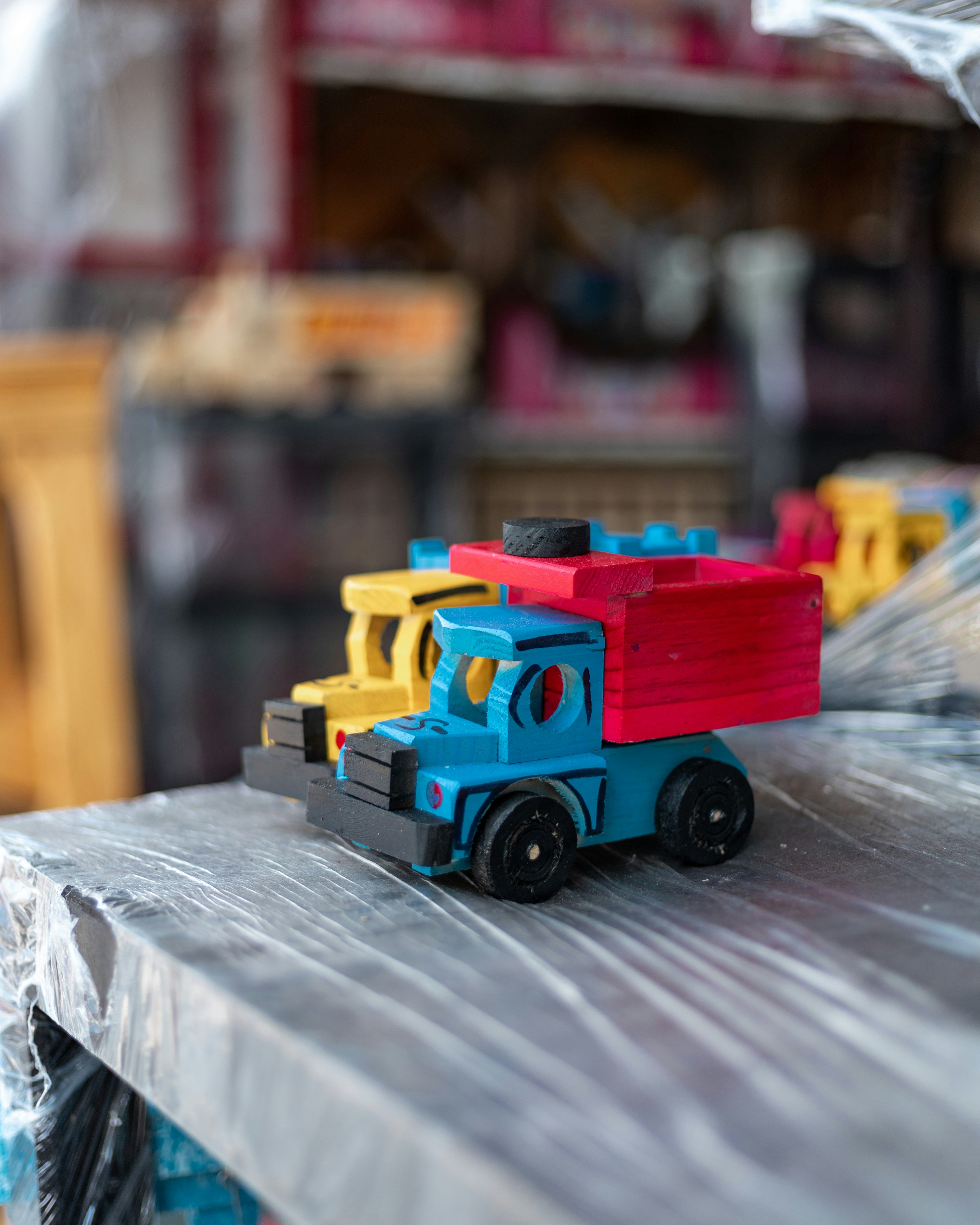 Colorful wooden toy trucks with red and blue paint sit on a shelf wrapped in plastic.