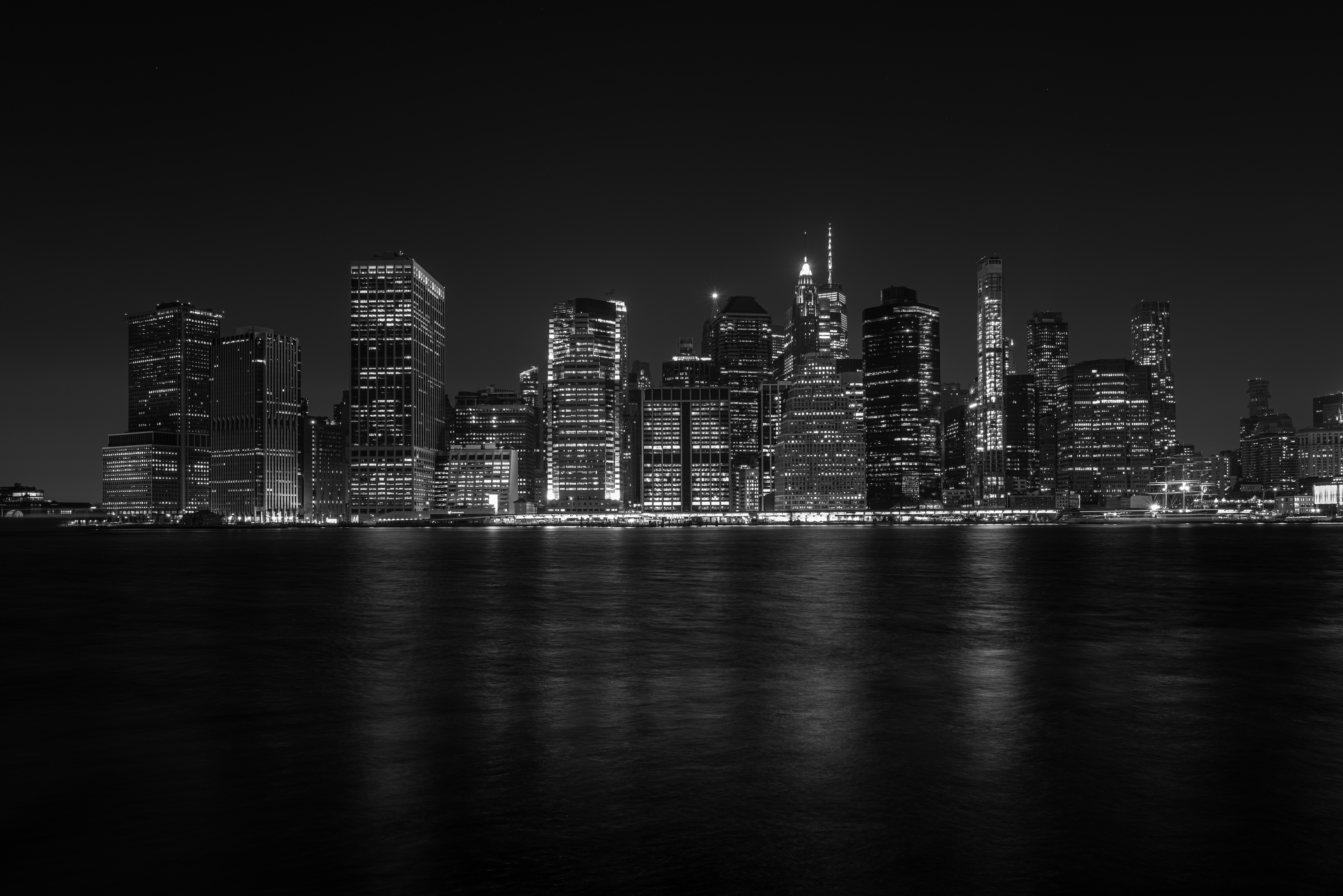 City skyline illuminated at night, showcasing a blend of modern architecture and reflective waters. A monochromatic palette emphasizes the contrast and textures of the buildings.