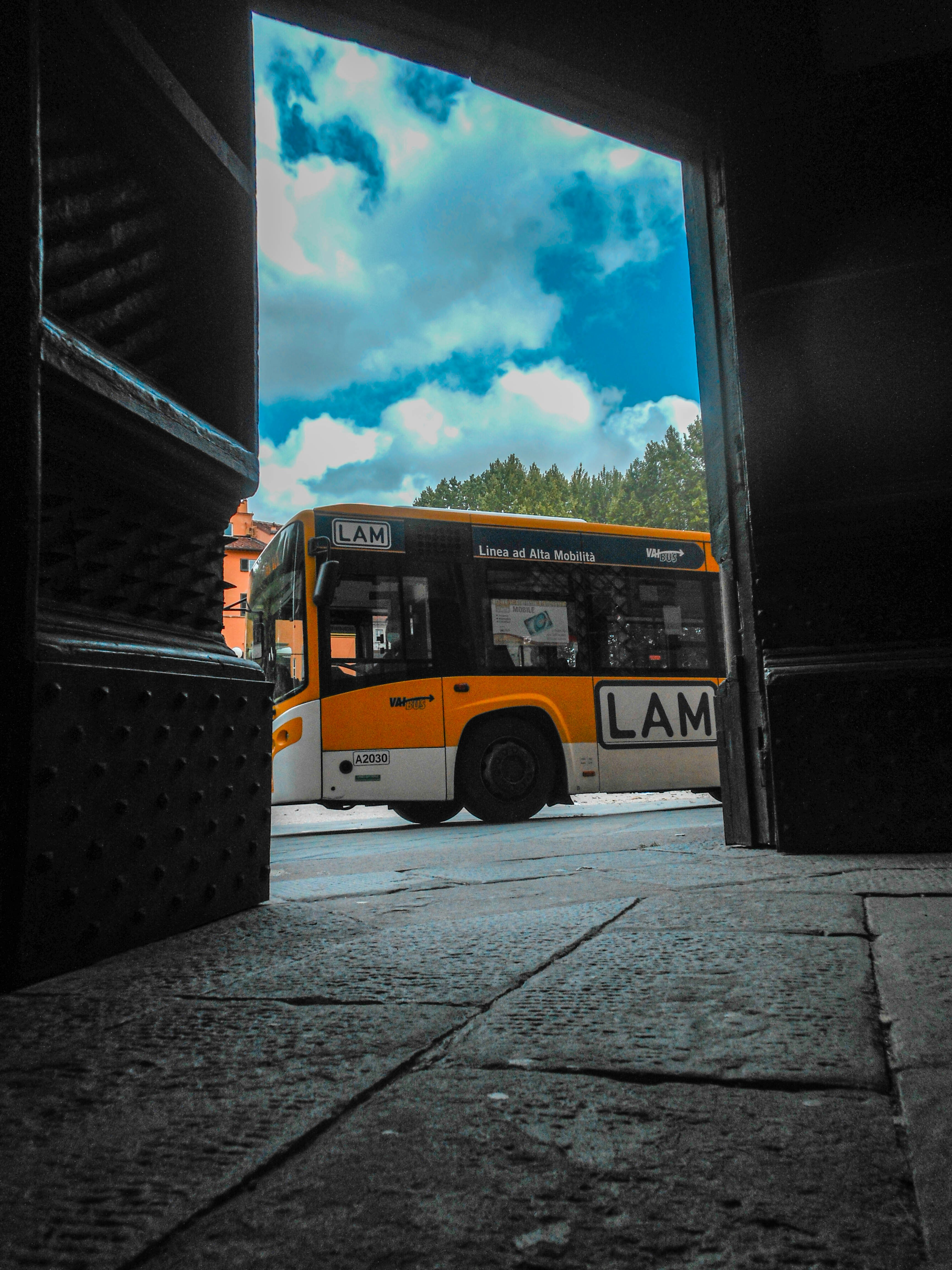 Yellow LAM bus framed by a dark stone arch, viewed from a low cobblestone foreground.