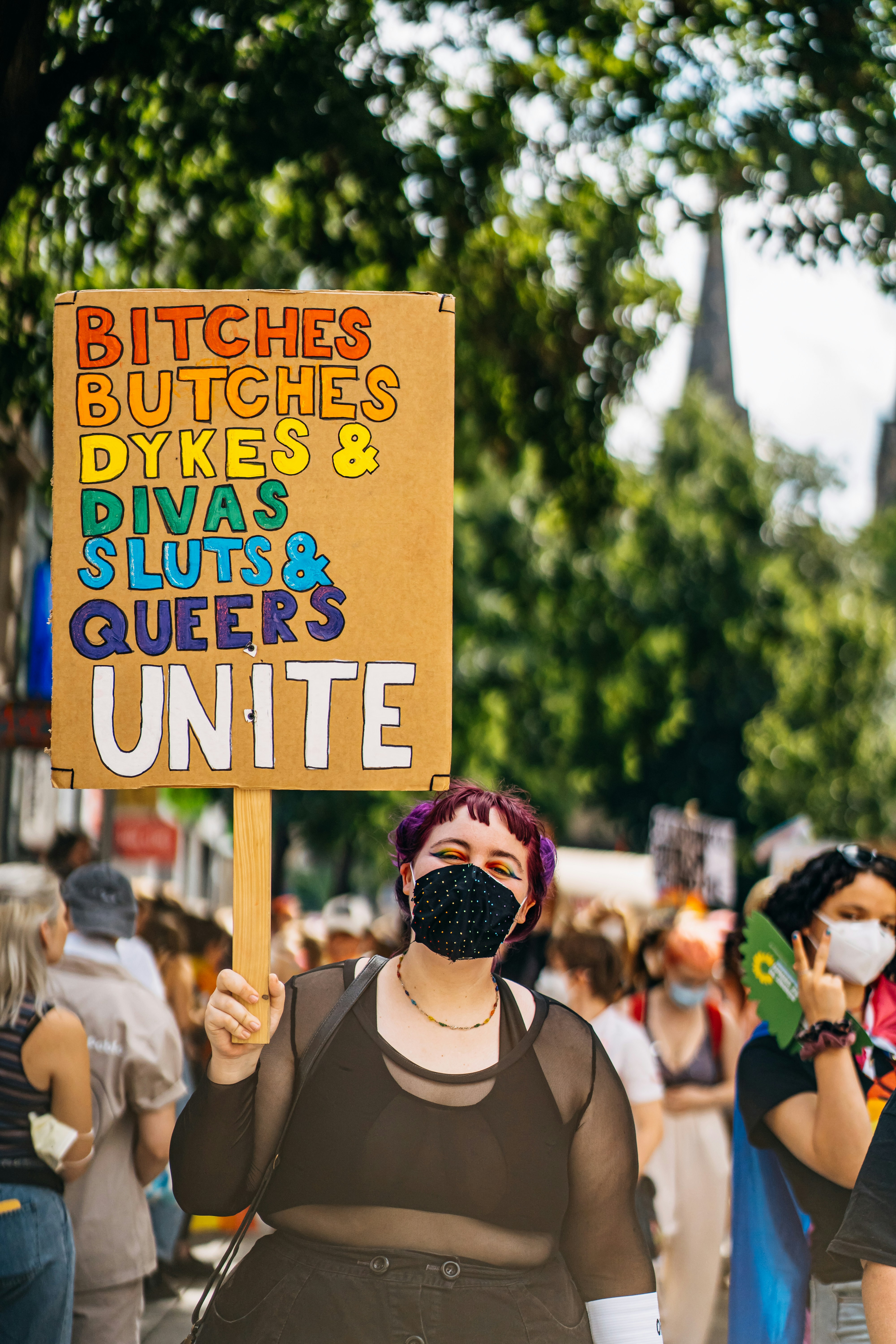 A person with purple hair holds a brightly colored sign advocating for LGBTQ+ solidarity at a vibrant rally. The crowd is blurred in the background, emphasizing the message of unity.