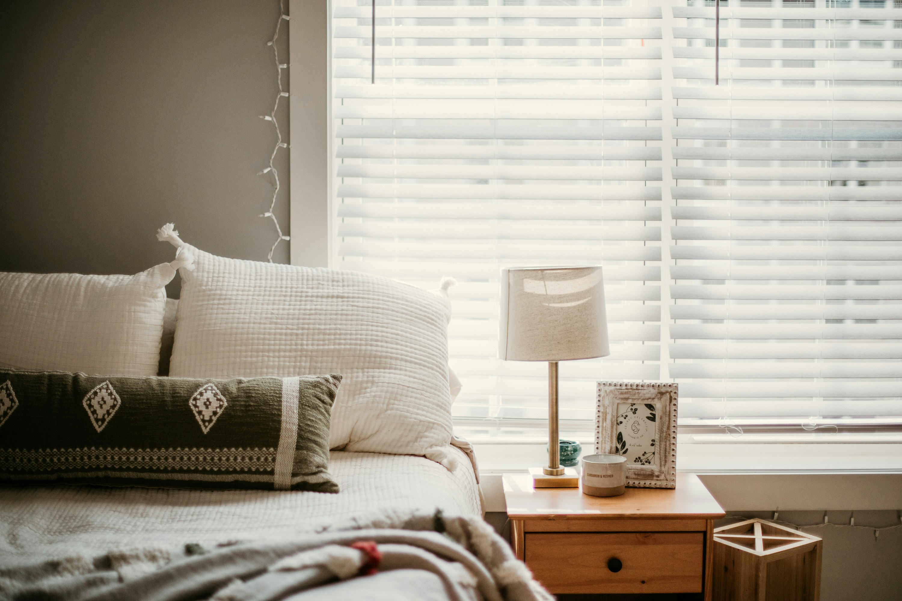 A minimalist bedroom featuring a neatly made bed with textured pillows, a bedside lamp, and decorative elements, illuminated by soft natural light from the window.