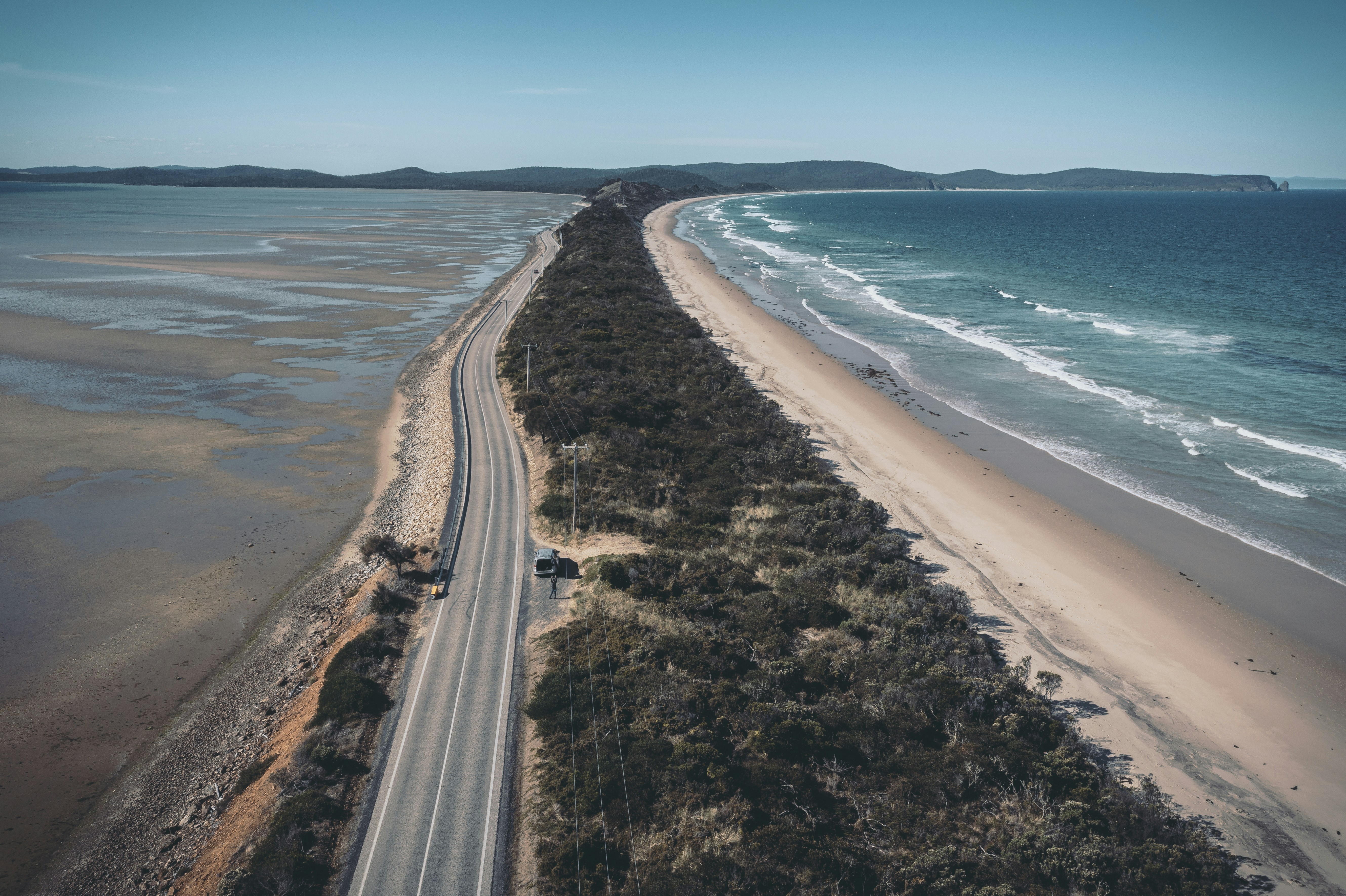 This striking aerial view captures a narrow road flanked by contrasting landscapes—a serene beach with turquoise waves on one side and a tranquil, expansive flatland on the other. The composition leads the eye along the road, emphasizing the dramatic meeting of land and sea. The vibrant blues of the ocean blend seamlessly with the earthy tones of the shoreline, under a clear sky, creating a harmonious and captivating scene.