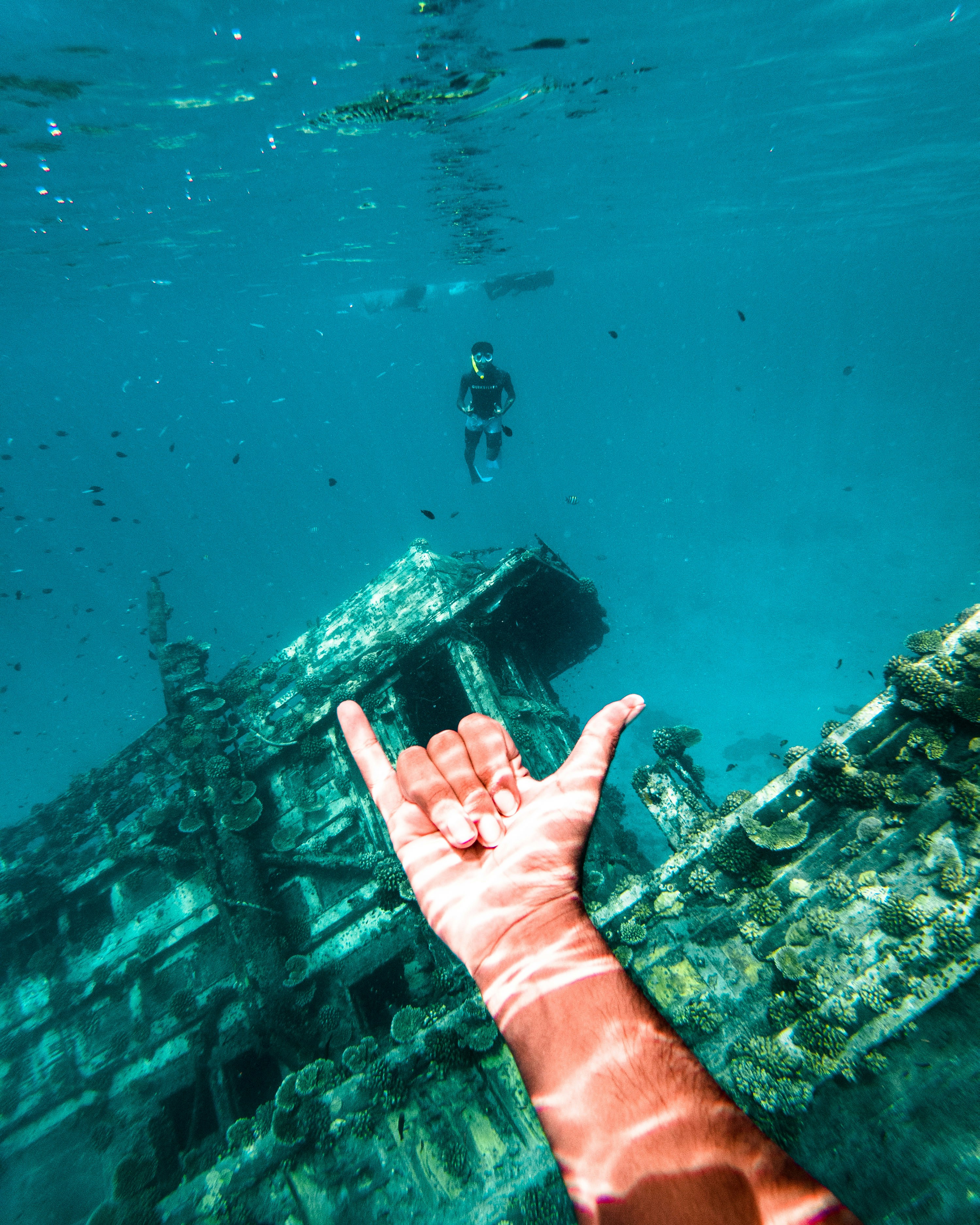 Underwater photograph of a diver hovering above a sunken wreck, with a large hand in the foreground making a rock-on gesture amid coral and marine life.