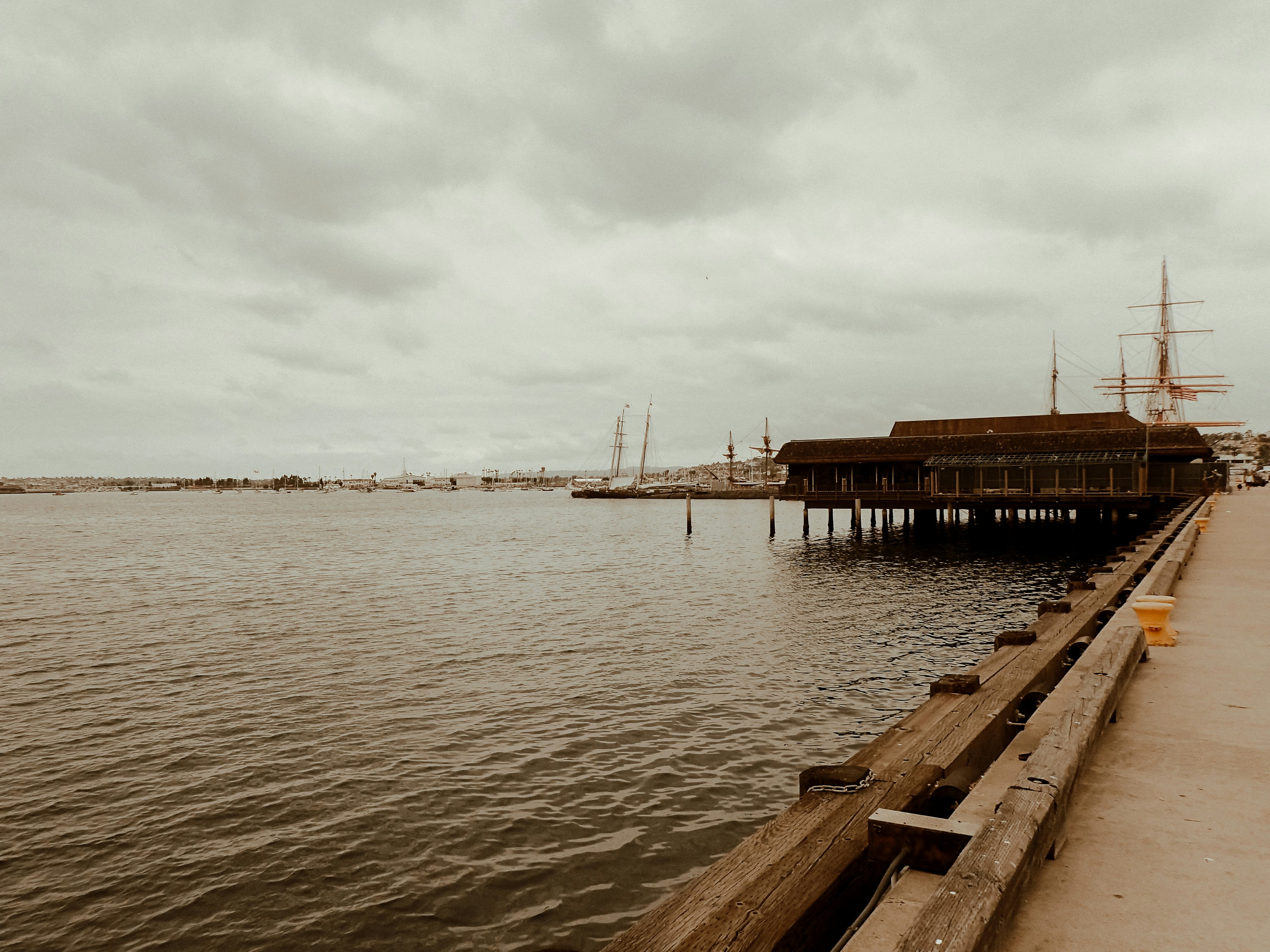 Wooden pier extending over calm waters, framed by historical ships and a cloudy sky.