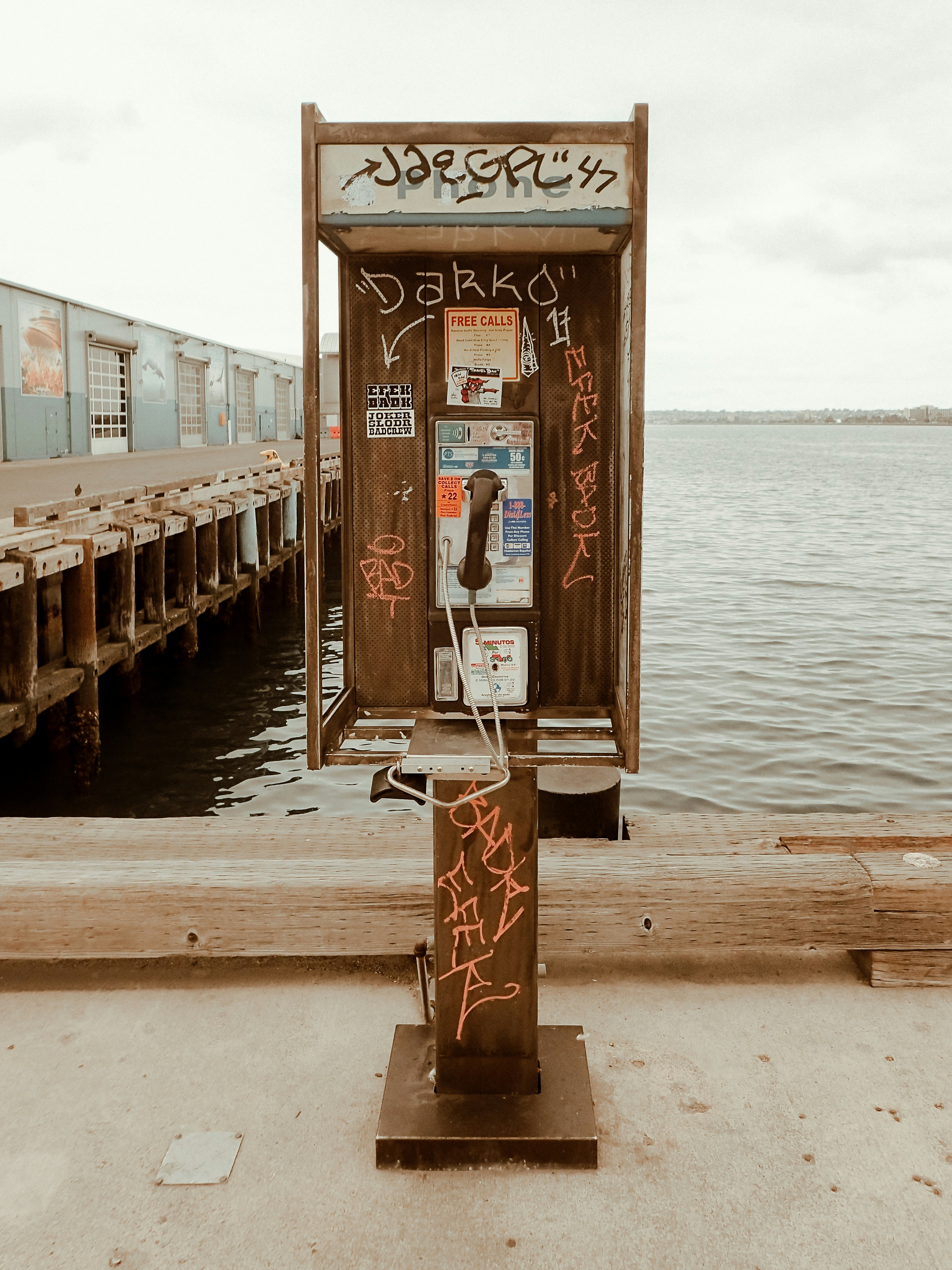Weathered payphone adorned with graffiti stands on a pier, overlooking a serene body of water. The juxtaposition of urban decay and tranquil scenery creates a poignant scene.