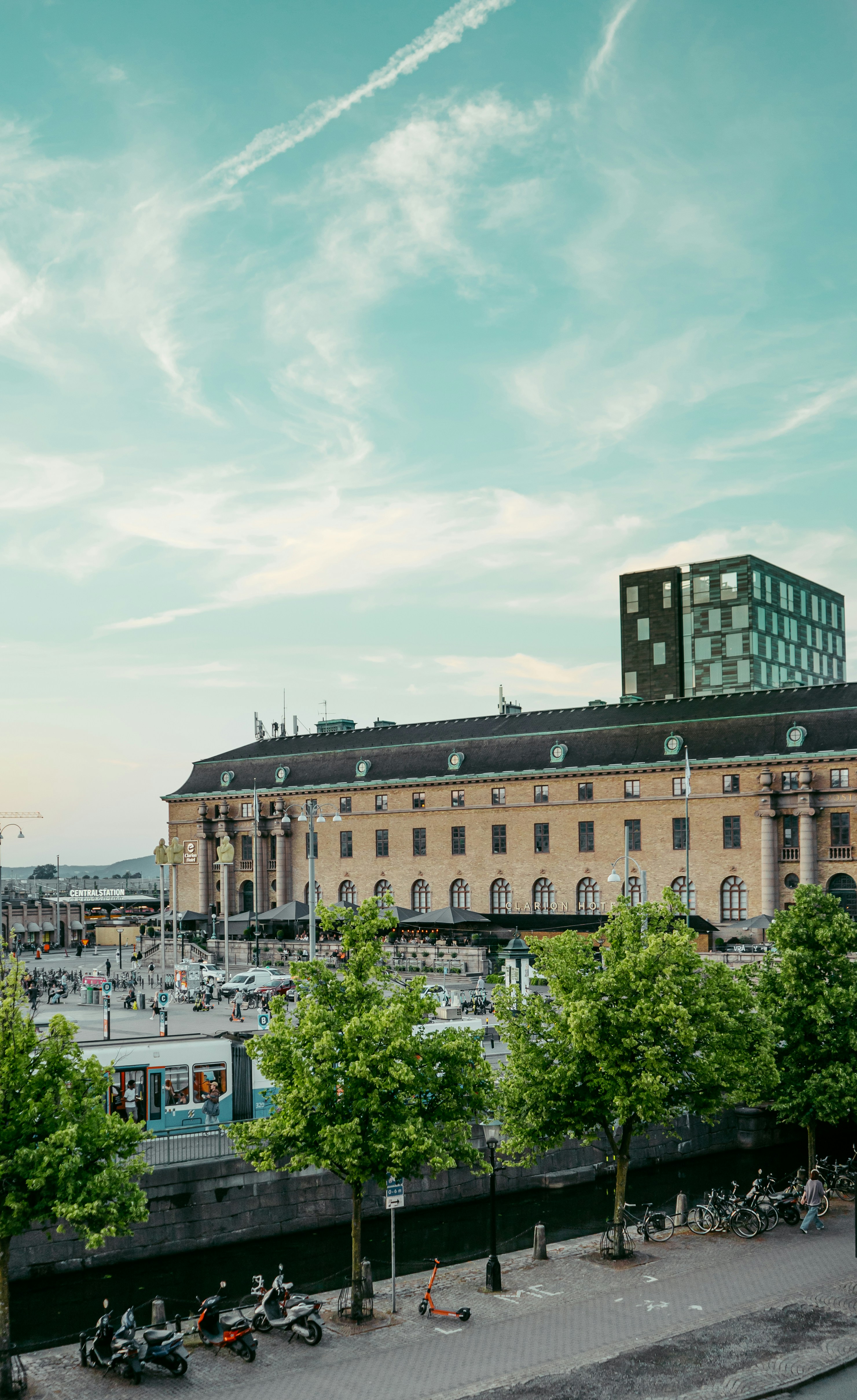 Historic building juxtaposed with modern architecture, framed by vibrant greenery and bustling urban life.
