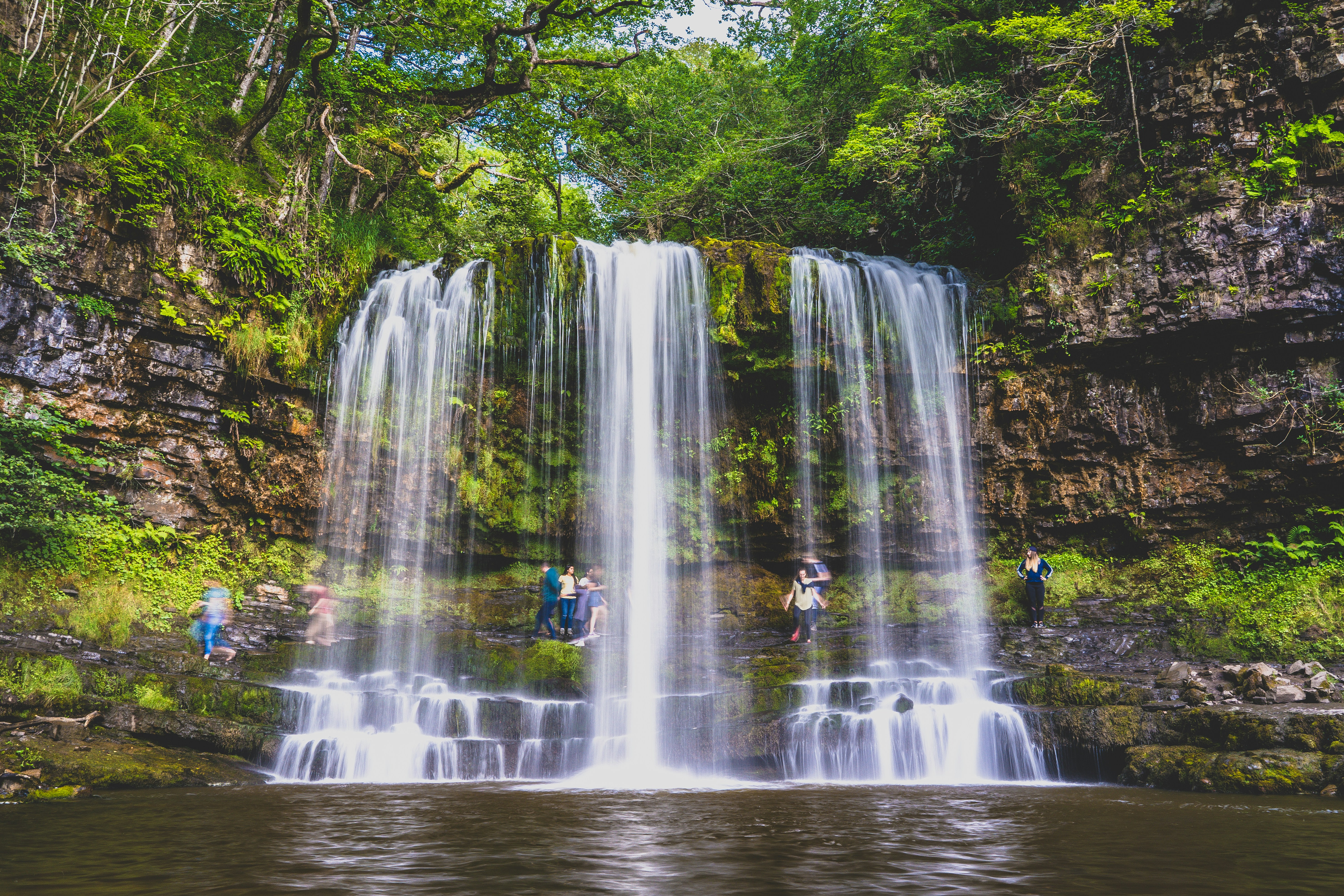 Waterfall cascading over a rocky cliff surrounded by lush greenery, with people exploring the scene.
