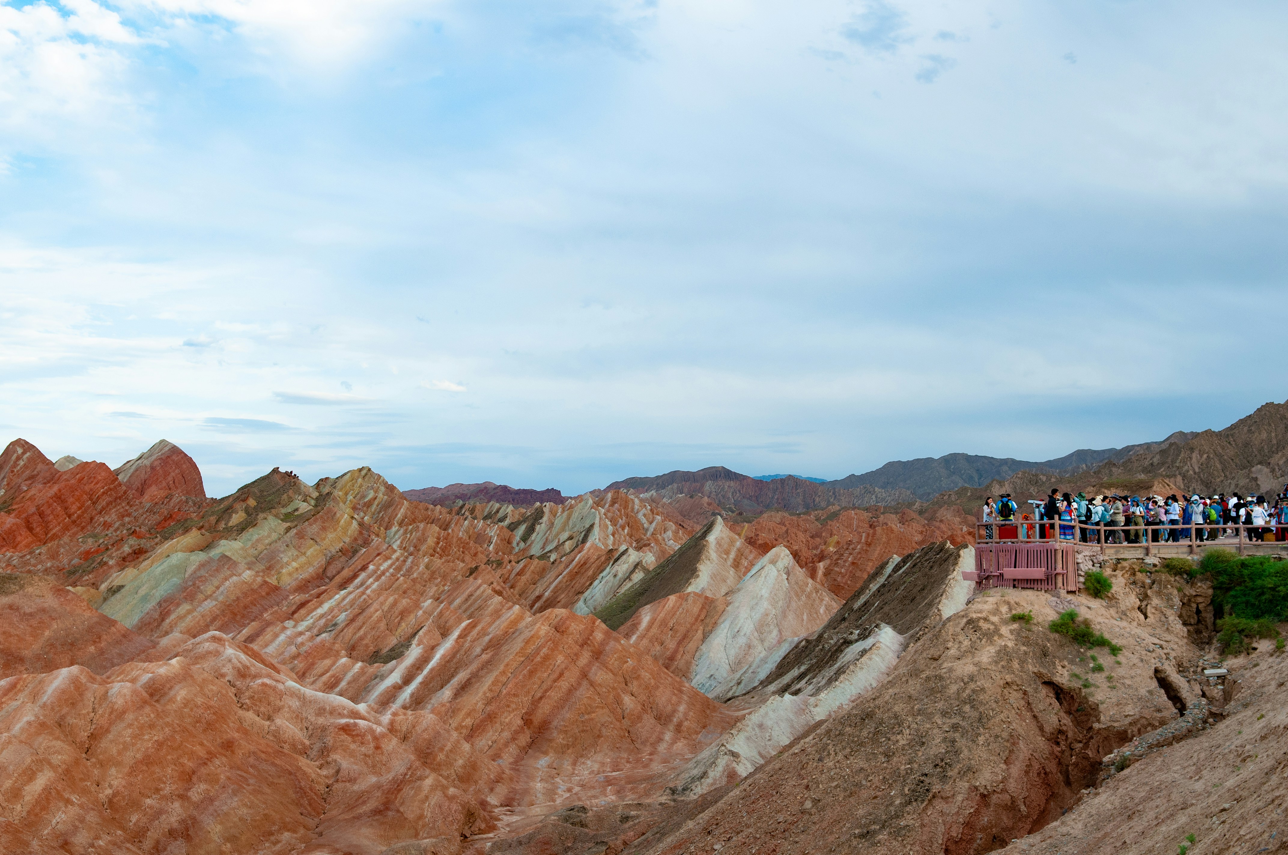 Vibrant geological formations stretch beneath a wide, clouded sky with visitors observing from a viewpoint.