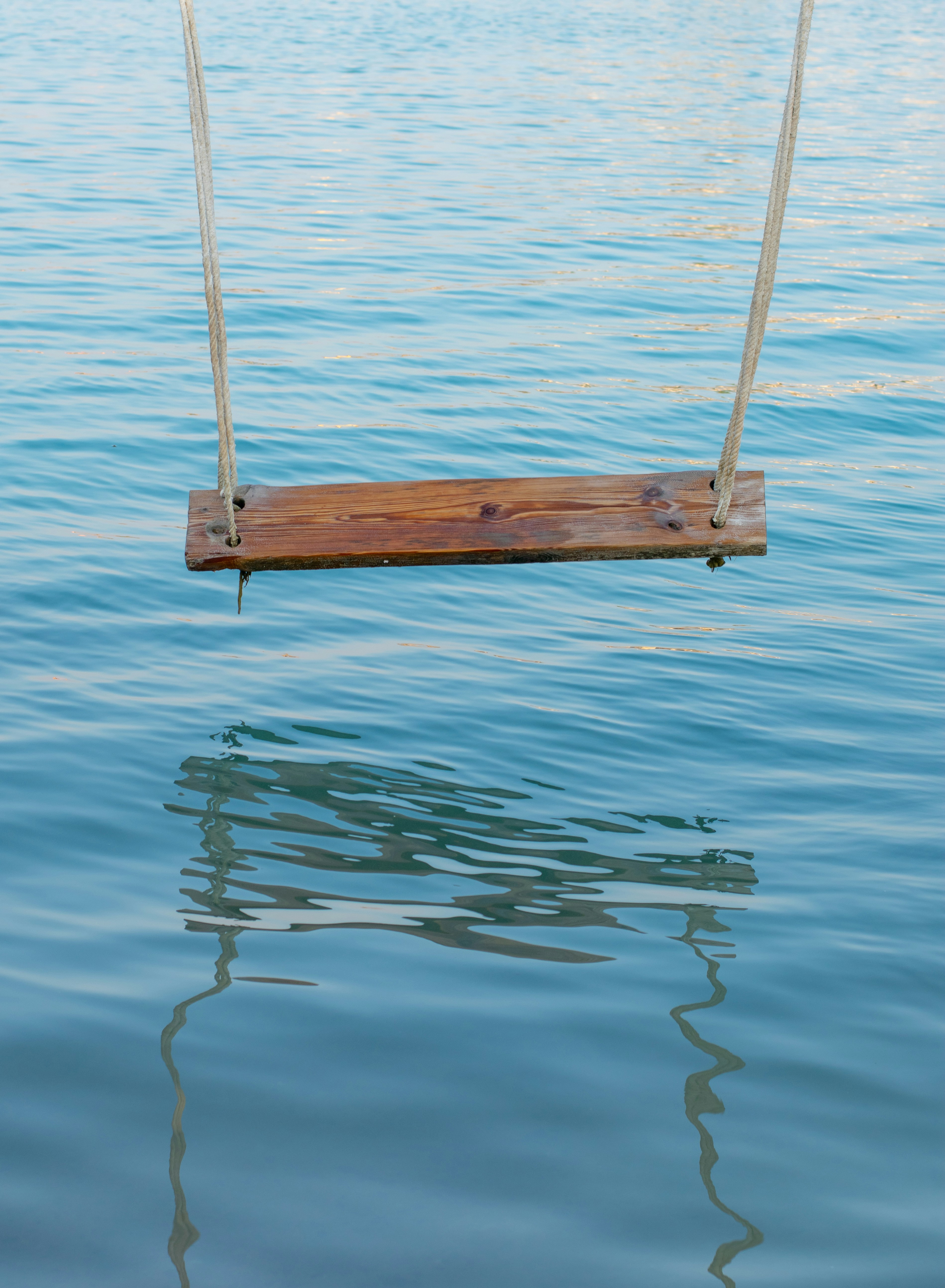 Wooden swing hangs above calm blue waters, reflecting its silhouette on the surface below.