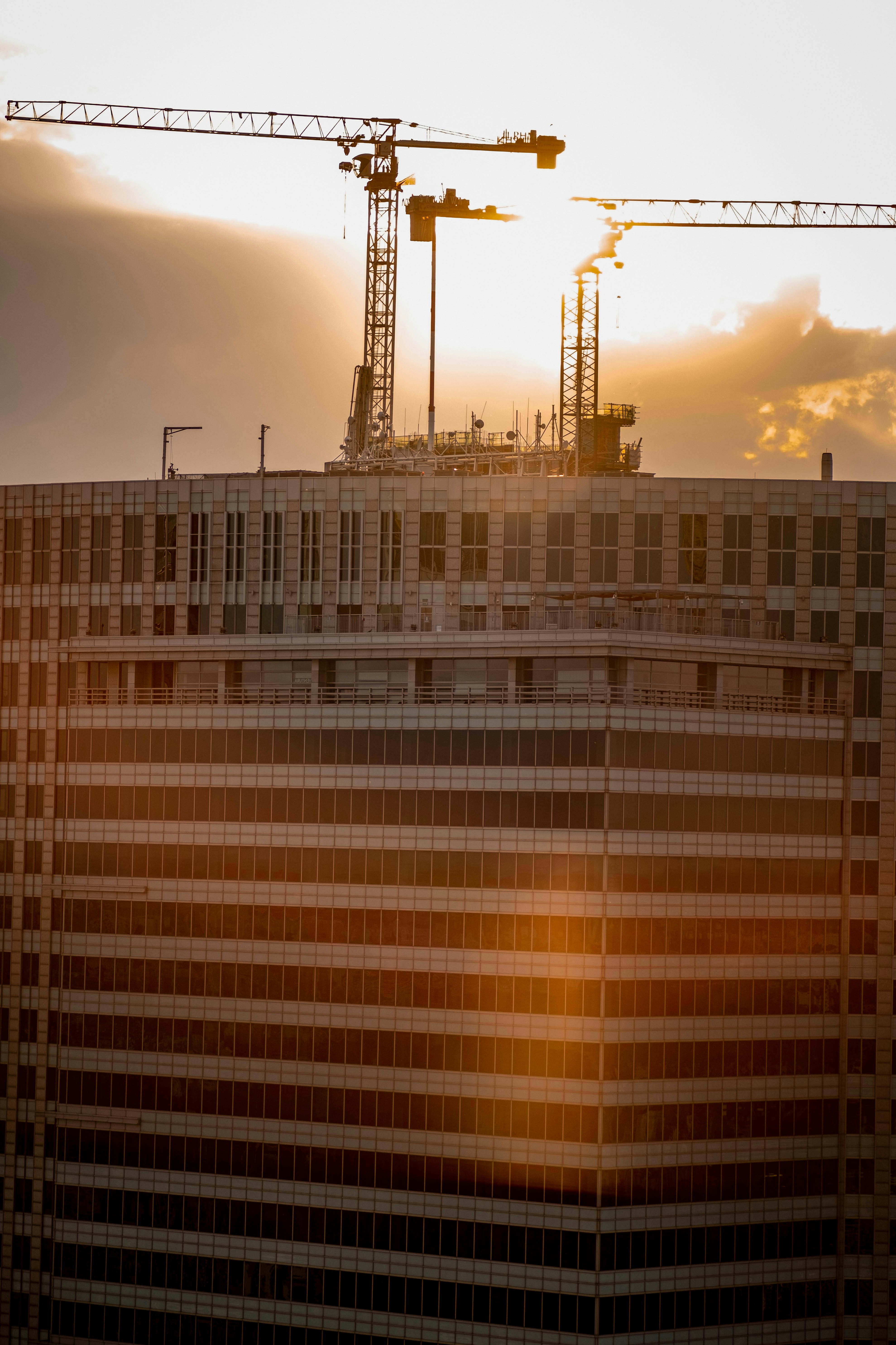 Construction cranes atop a modern building silhouetted against a vibrant sunset sky.