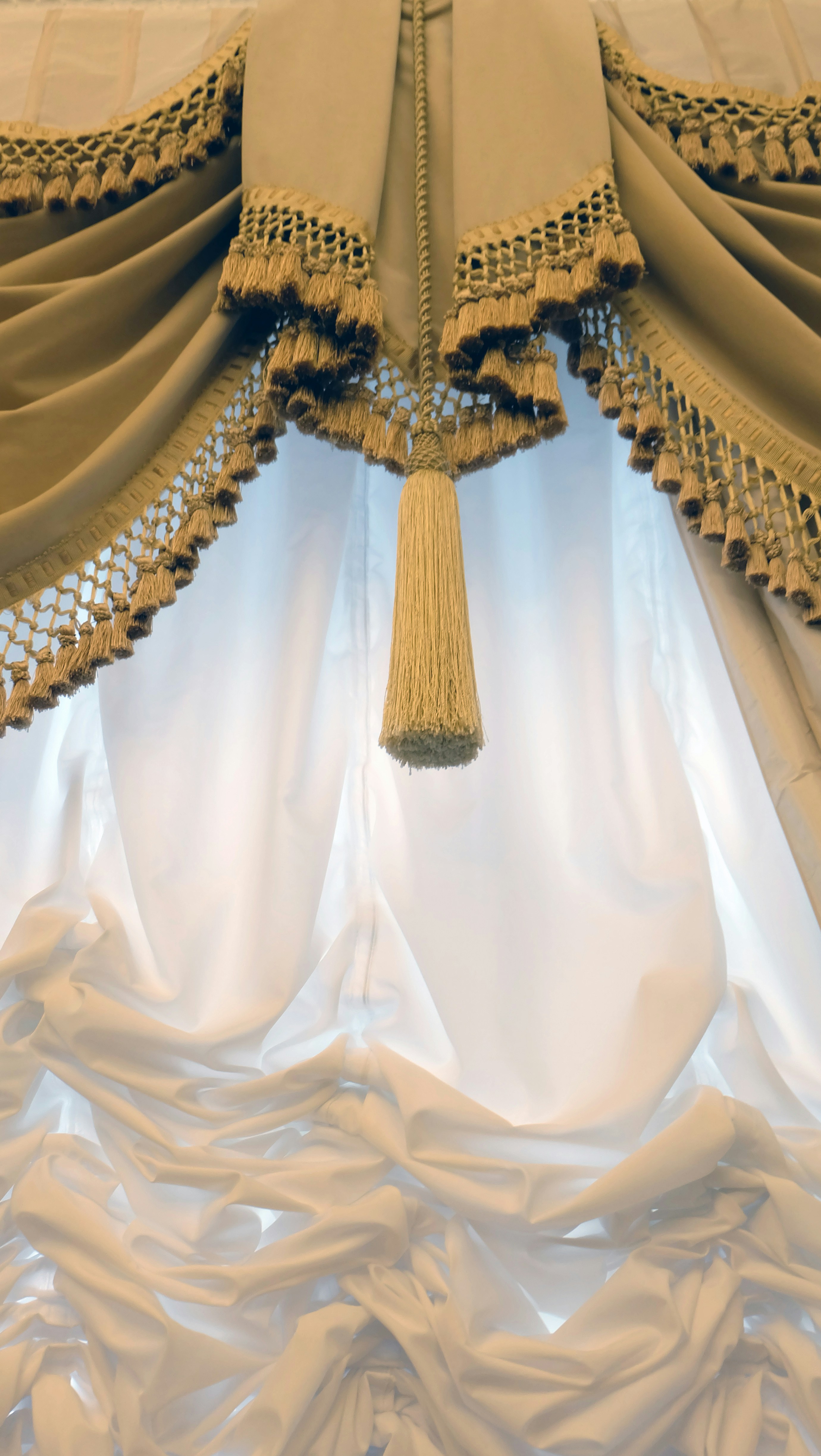 Intricately designed curtain draped elegantly, with a decorative tassel and soft white fabric behind it. The interplay of light creates a serene atmosphere.