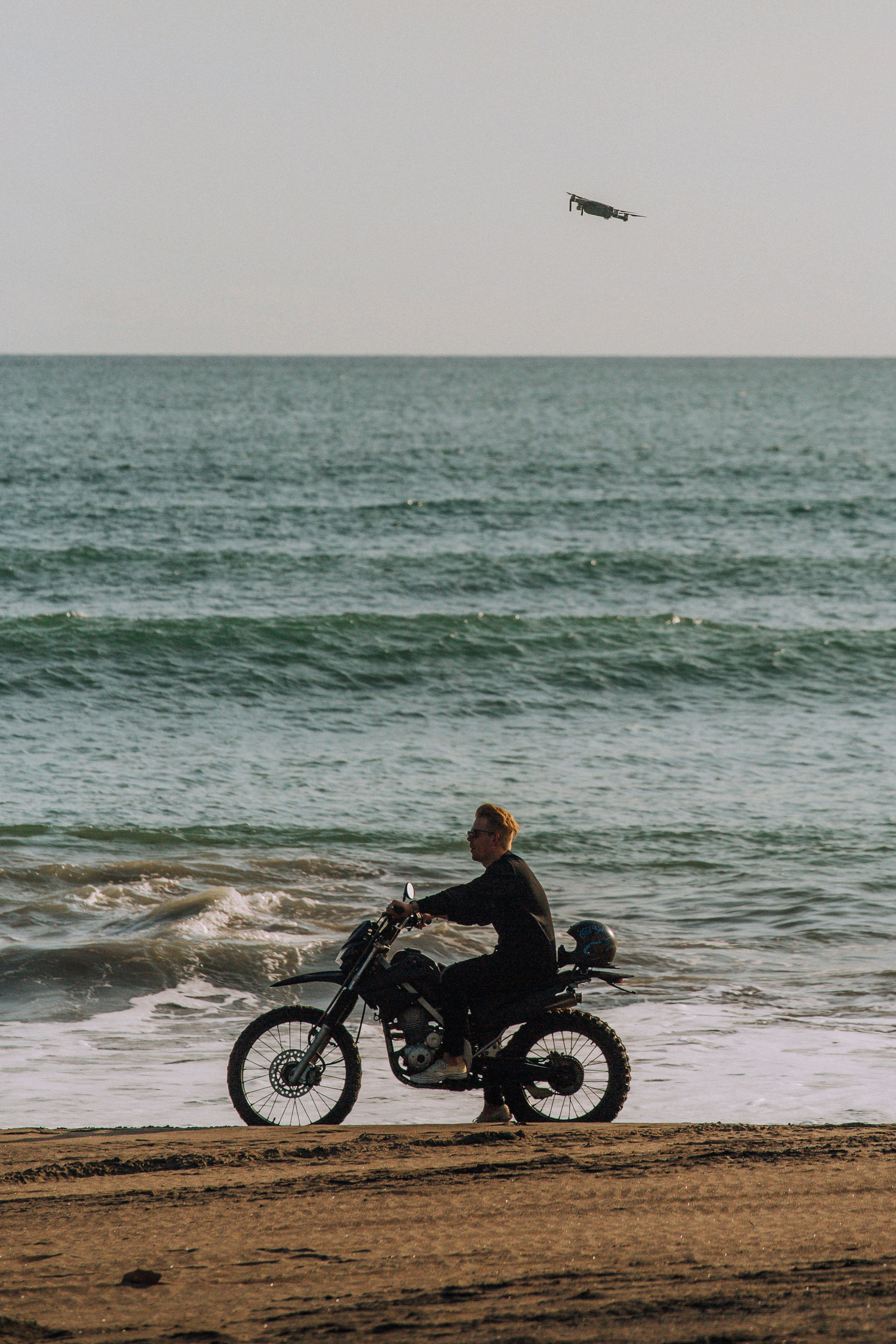 A motorcyclist pauses on the beach, gazing at the waves while a drone hovers above, illustrating the blend of technology and nature.