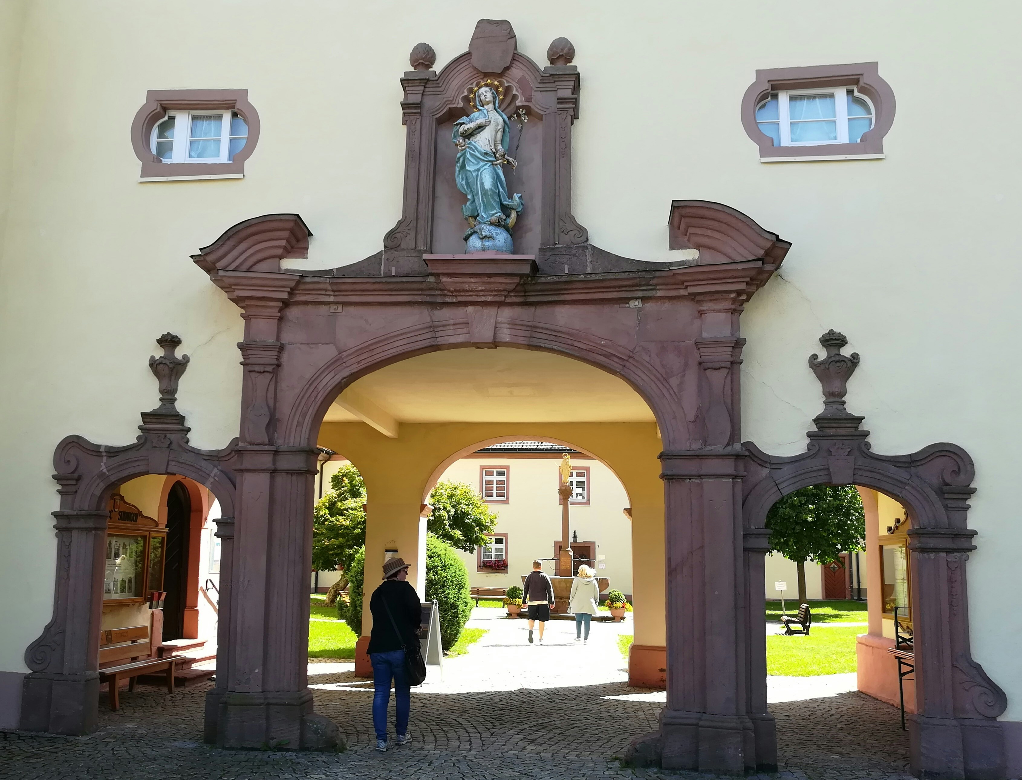 Ornate stone archway with a statue leads to a sunlit courtyard.