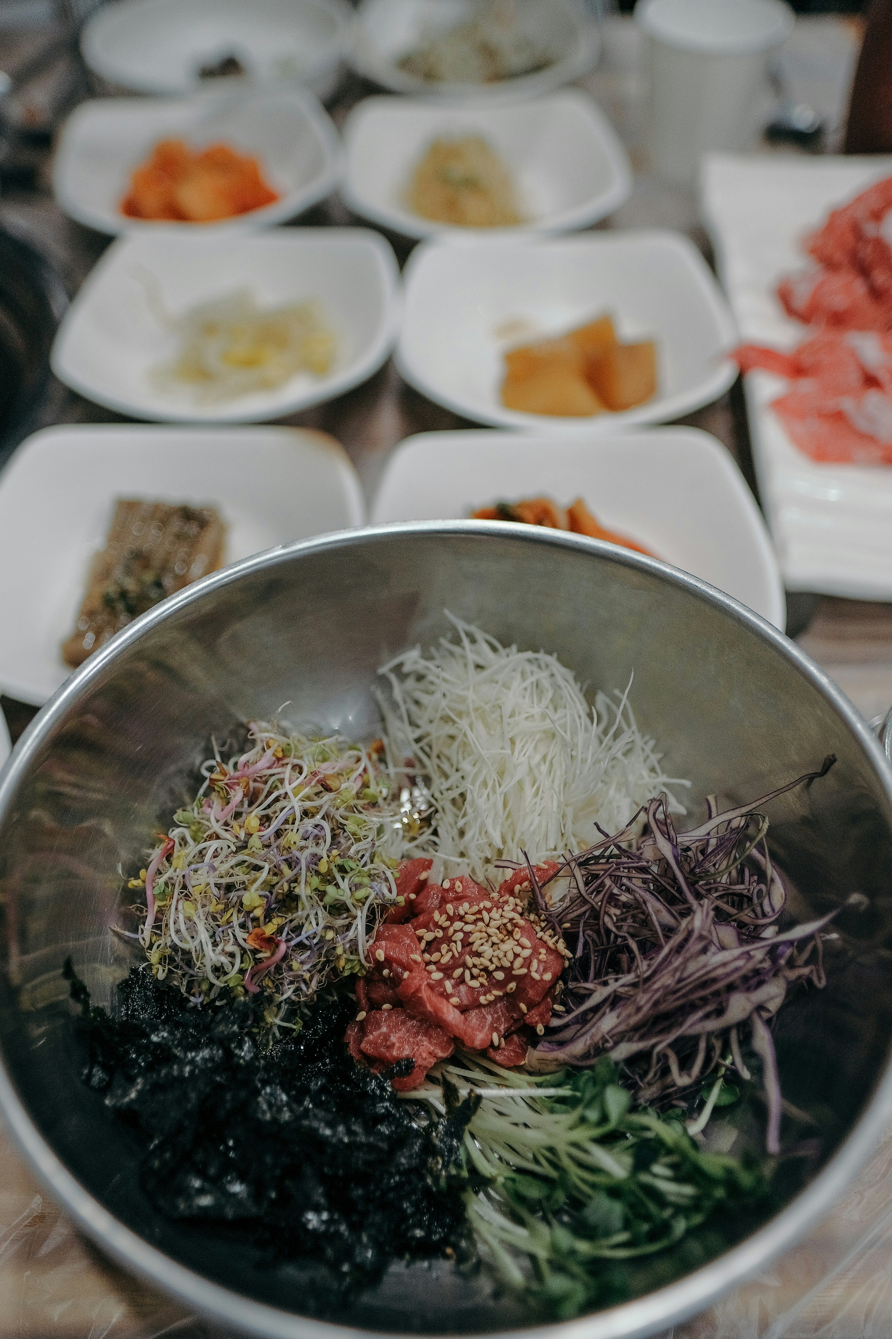 Colorful assortment of fresh ingredients in a metal bowl, surrounded by various side dishes on white plates. Perfectly arranged for a traditional Korean meal.