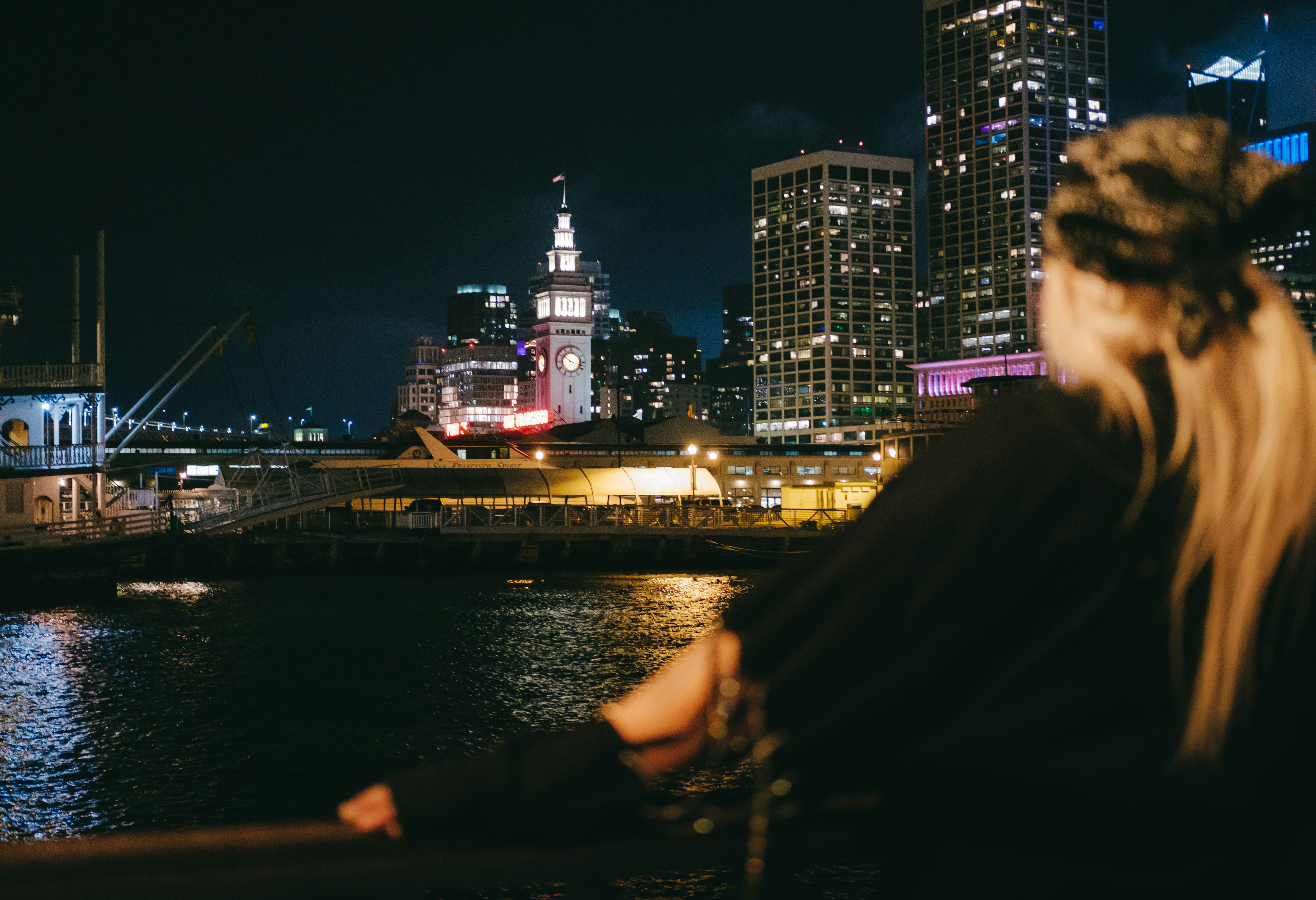 Person gazing over illuminated waterfront with city skyline in the background at night.