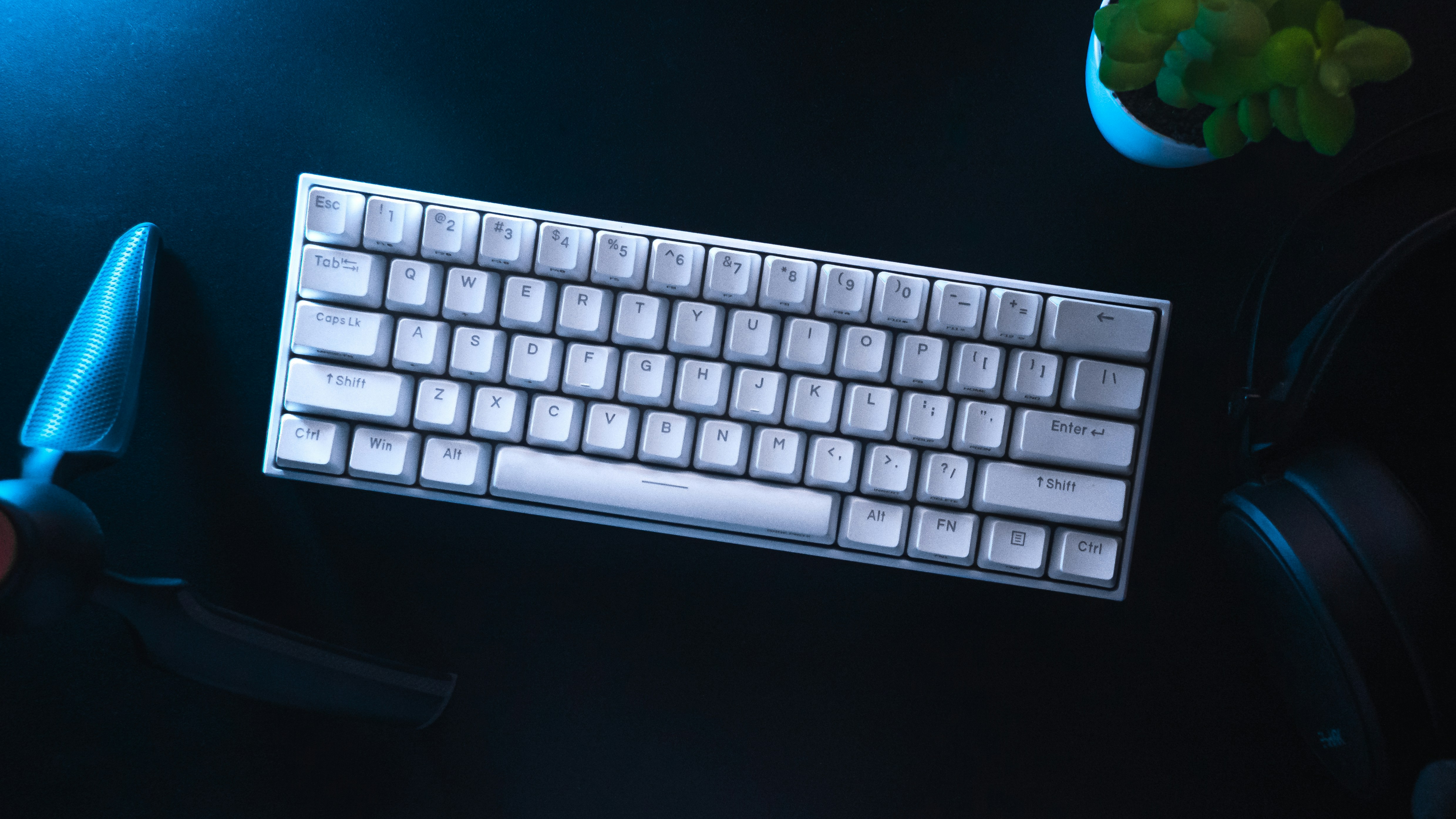 A sleek, white mechanical keyboard sits atop a dark surface, complemented by a potted plant and gaming headset, creating a modern workspace aesthetic.