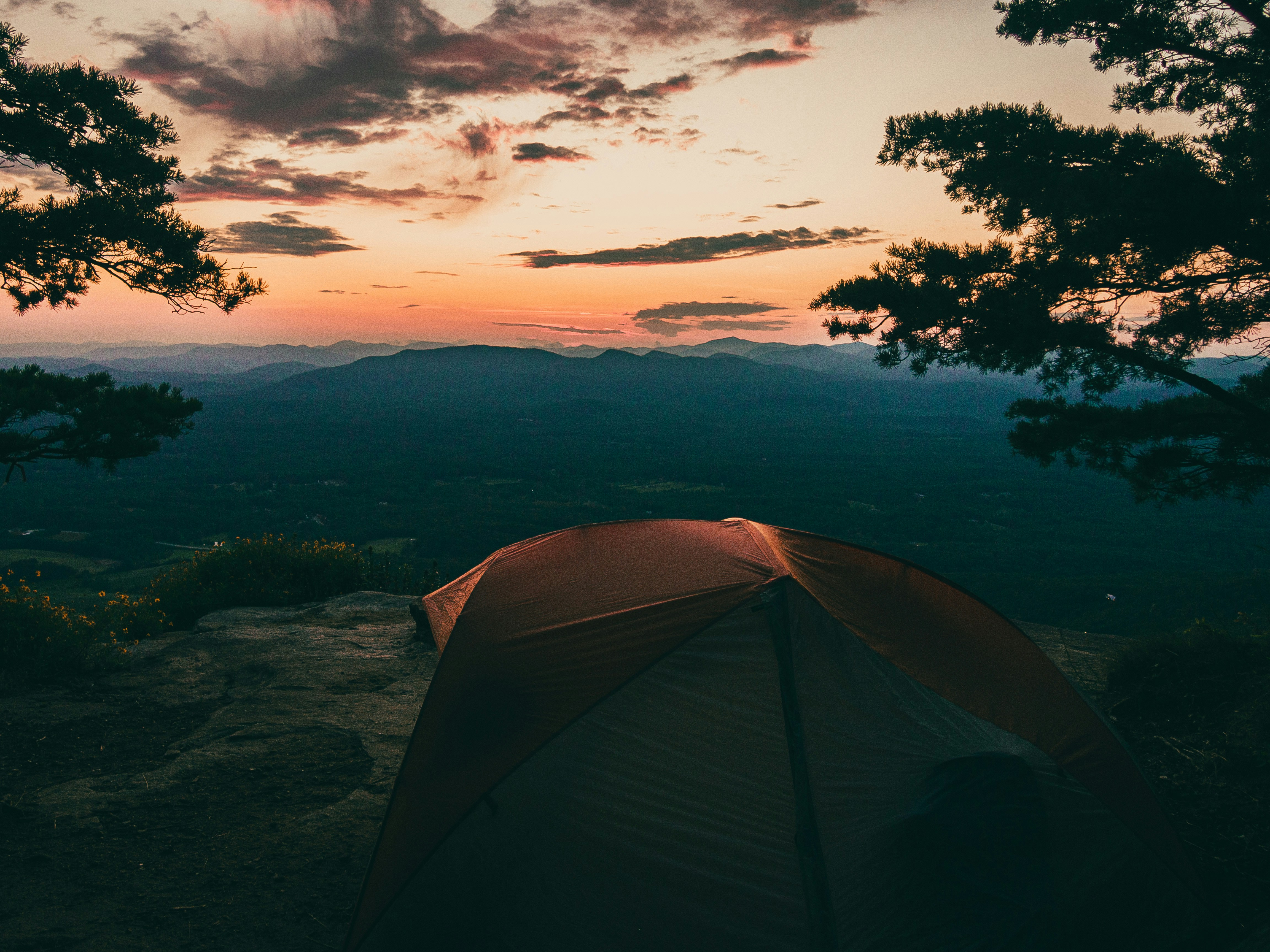 Tent pitched on a rocky outcrop overlooking a vast valley at sunset, framed by silhouetted trees. The warm hues of the sky contrast with the cool shadows of the landscape.