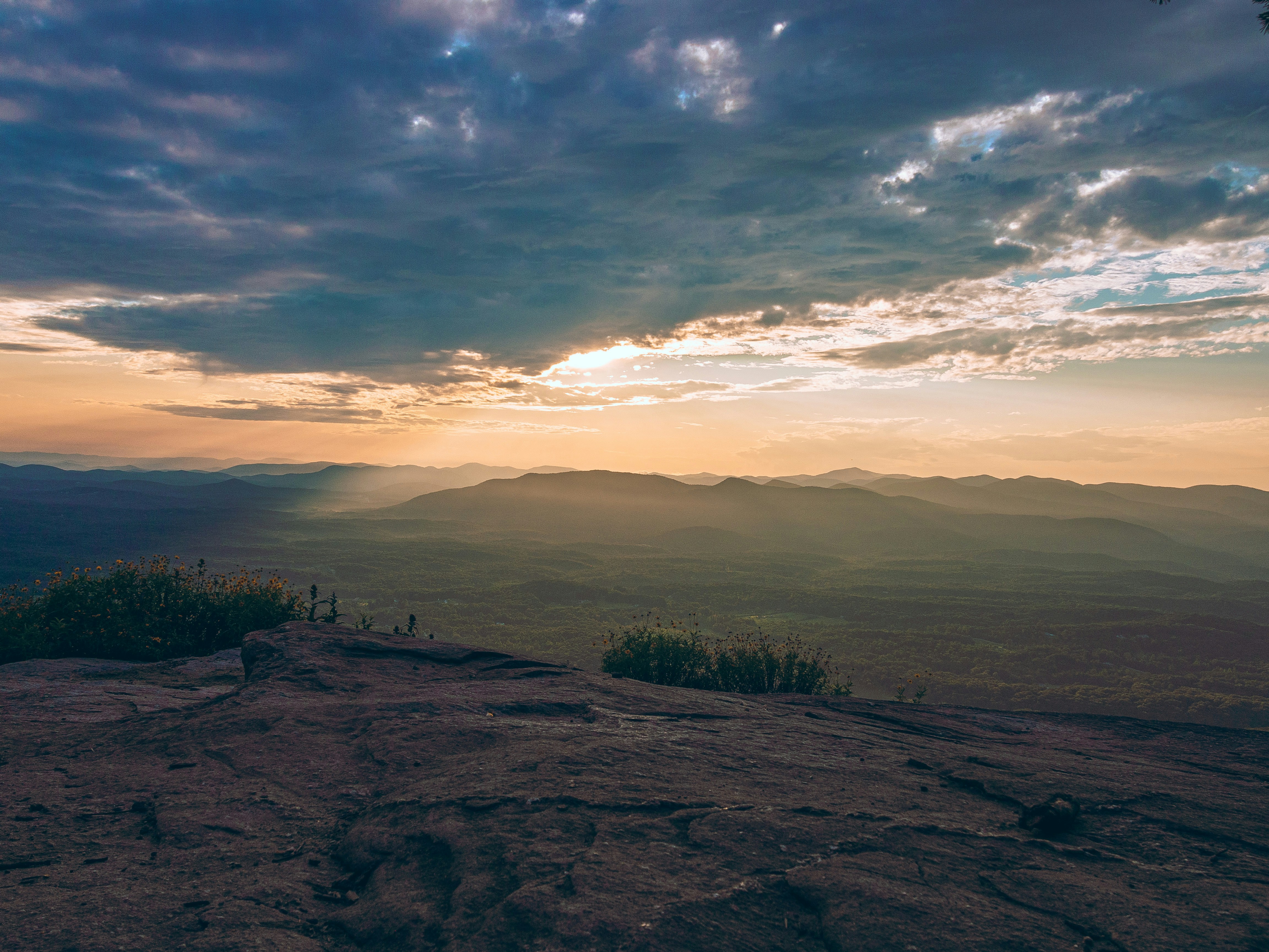 Golden sunlight filters through clouds, illuminating distant mountains and rocky foreground. The scene captures the tranquil transition of day to night.