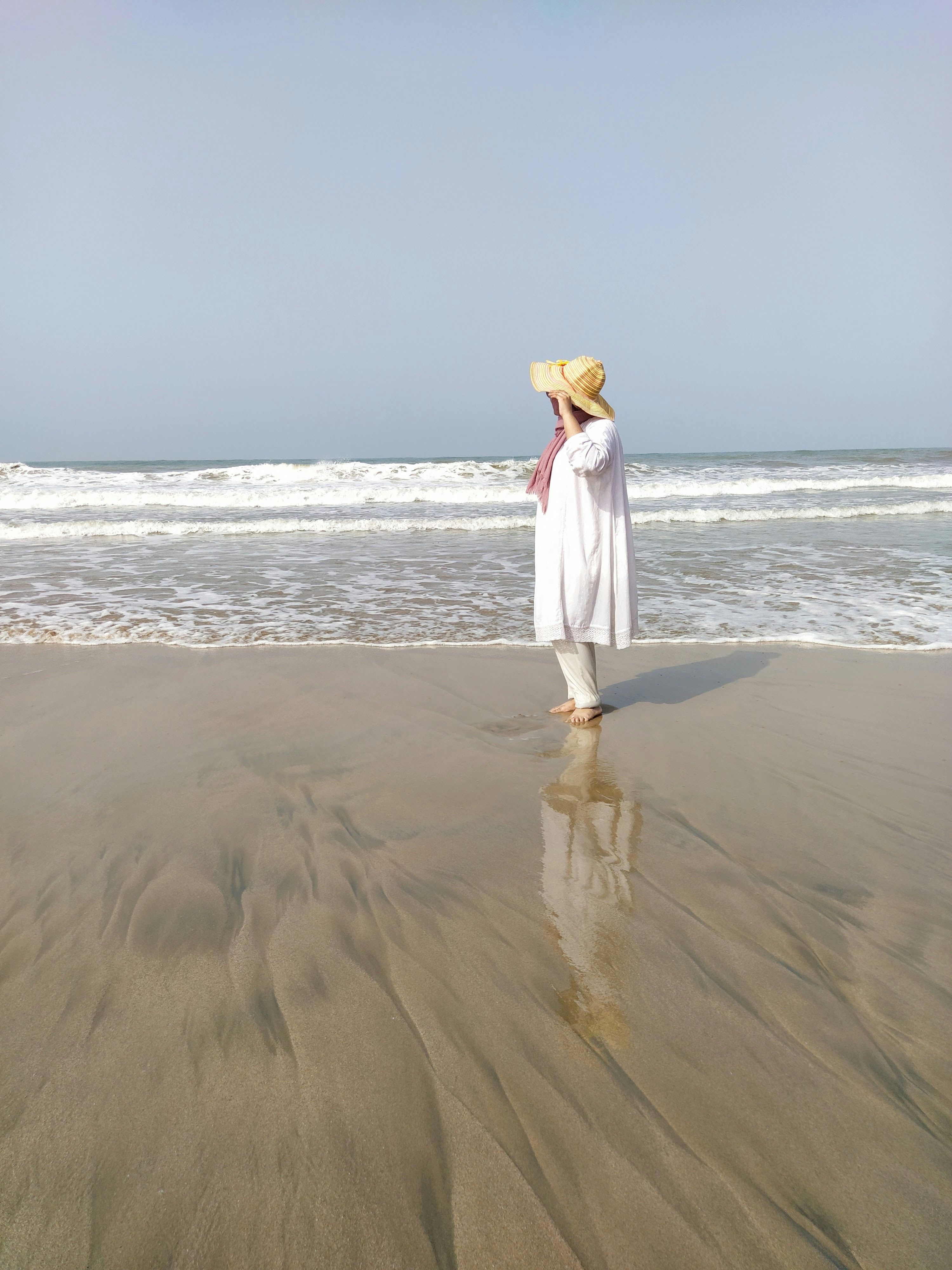 Photograph of a person in white with a straw hat standing on a wet beach as waves roll in. Their reflection stretches across the sand.