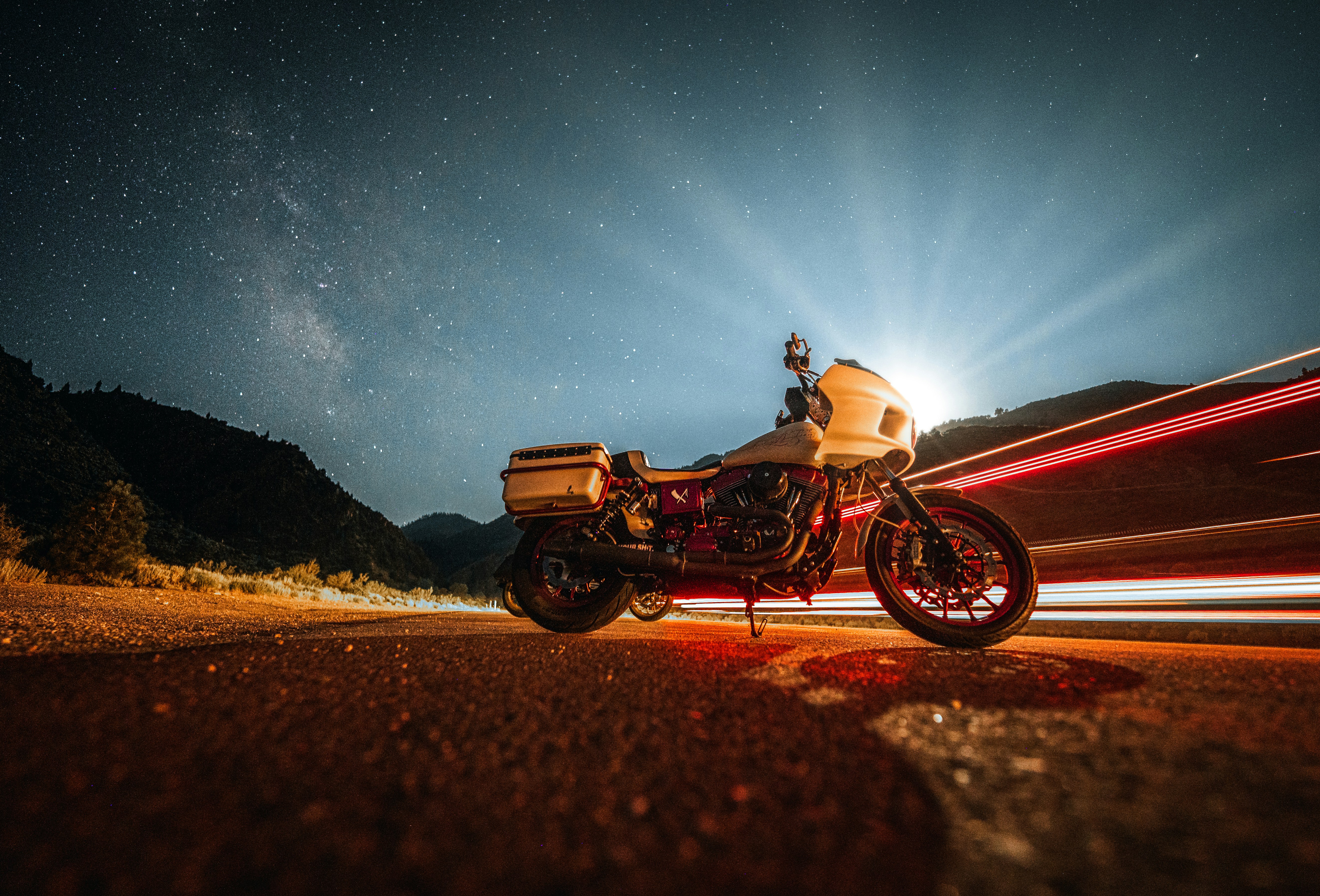 Motorcycle parked on a winding road illuminated by moonlight, with starry sky and light trails from passing vehicles in the background.