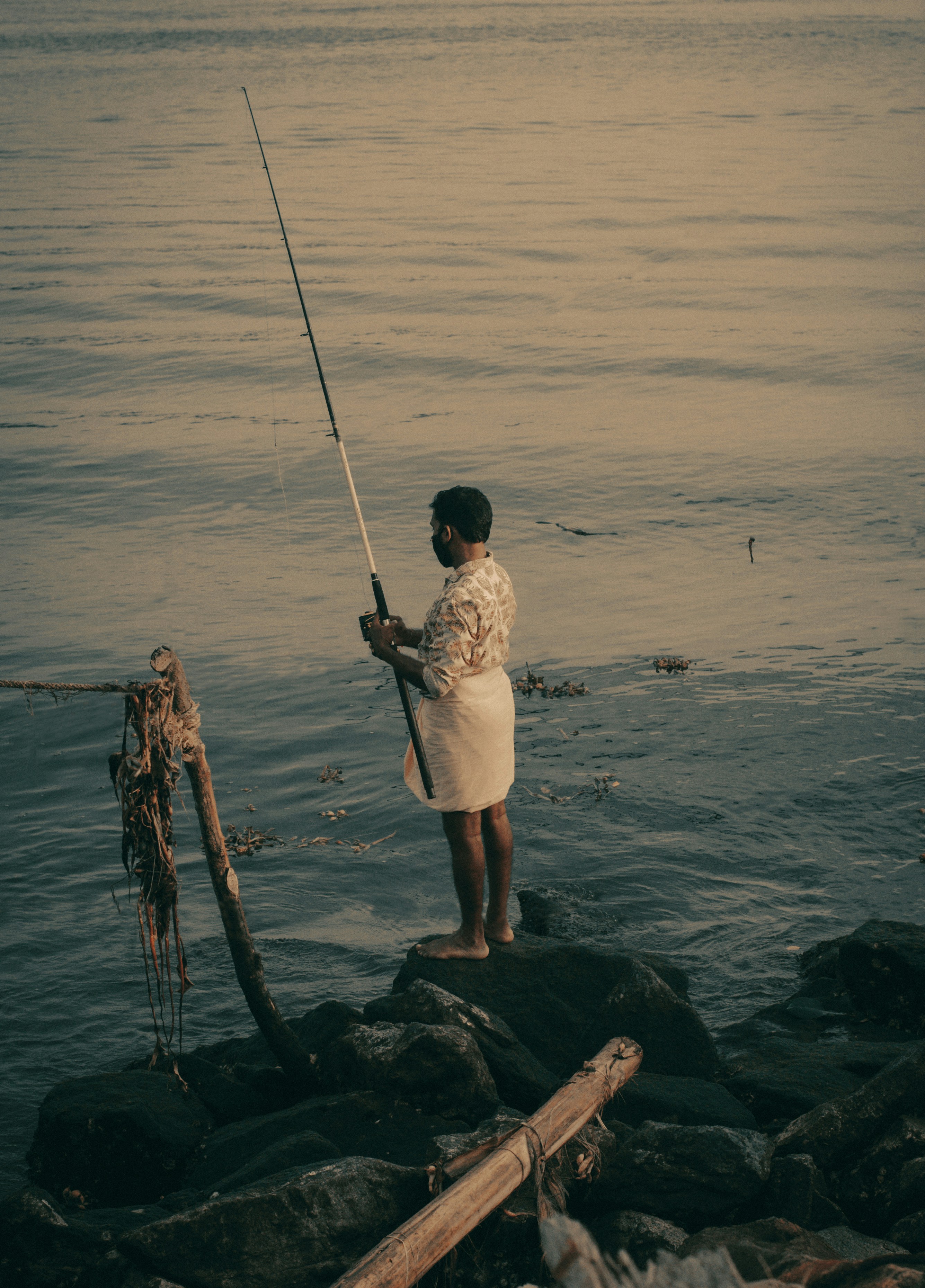 Fisherman standing on rocky shore, casting line into calm waters during golden hour. Soft light enhances the tranquil atmosphere.