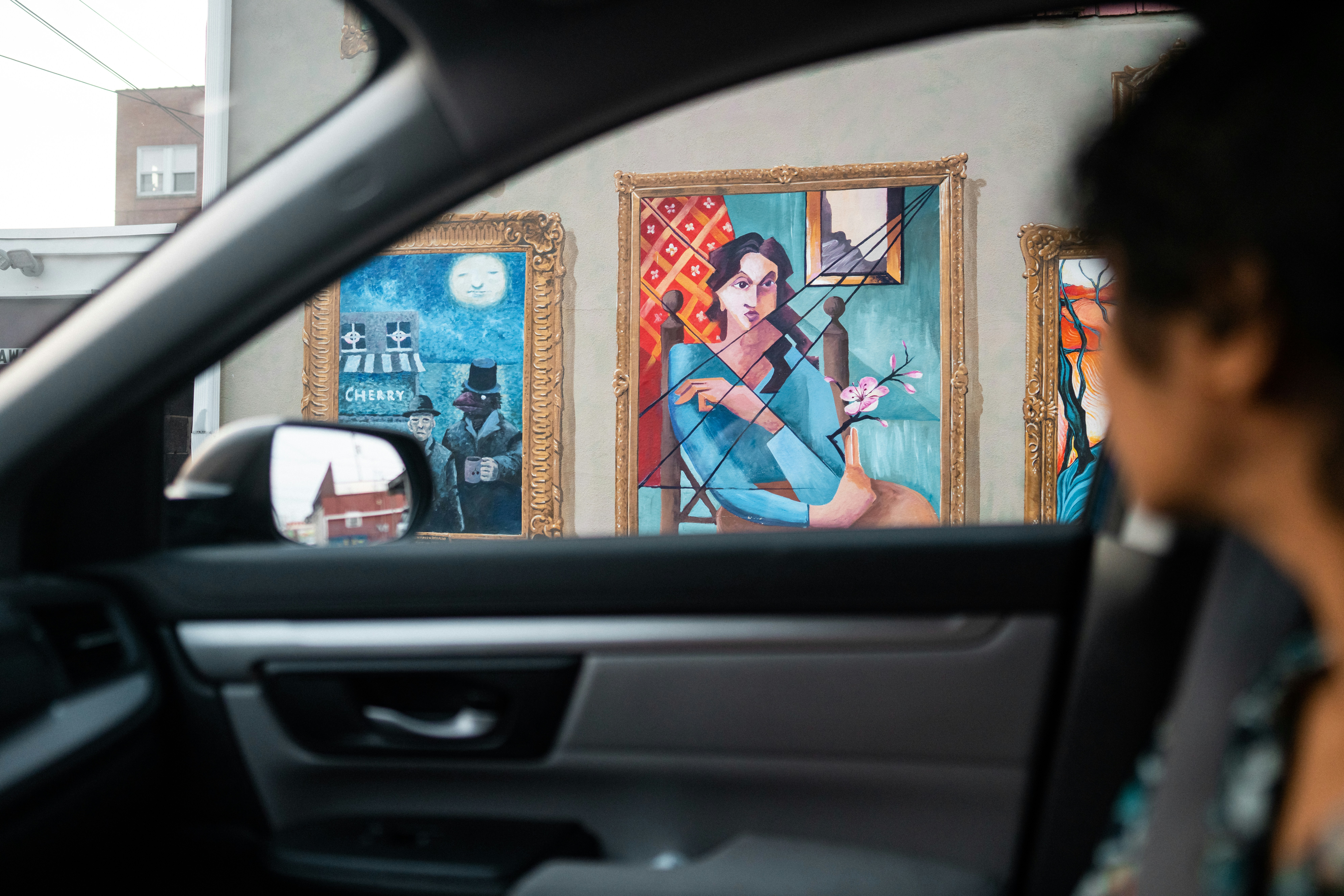 Vibrant artwork displayed in ornate frames, viewed from inside a car, with a person partially visible. The scene captures the intersection of art and daily life.