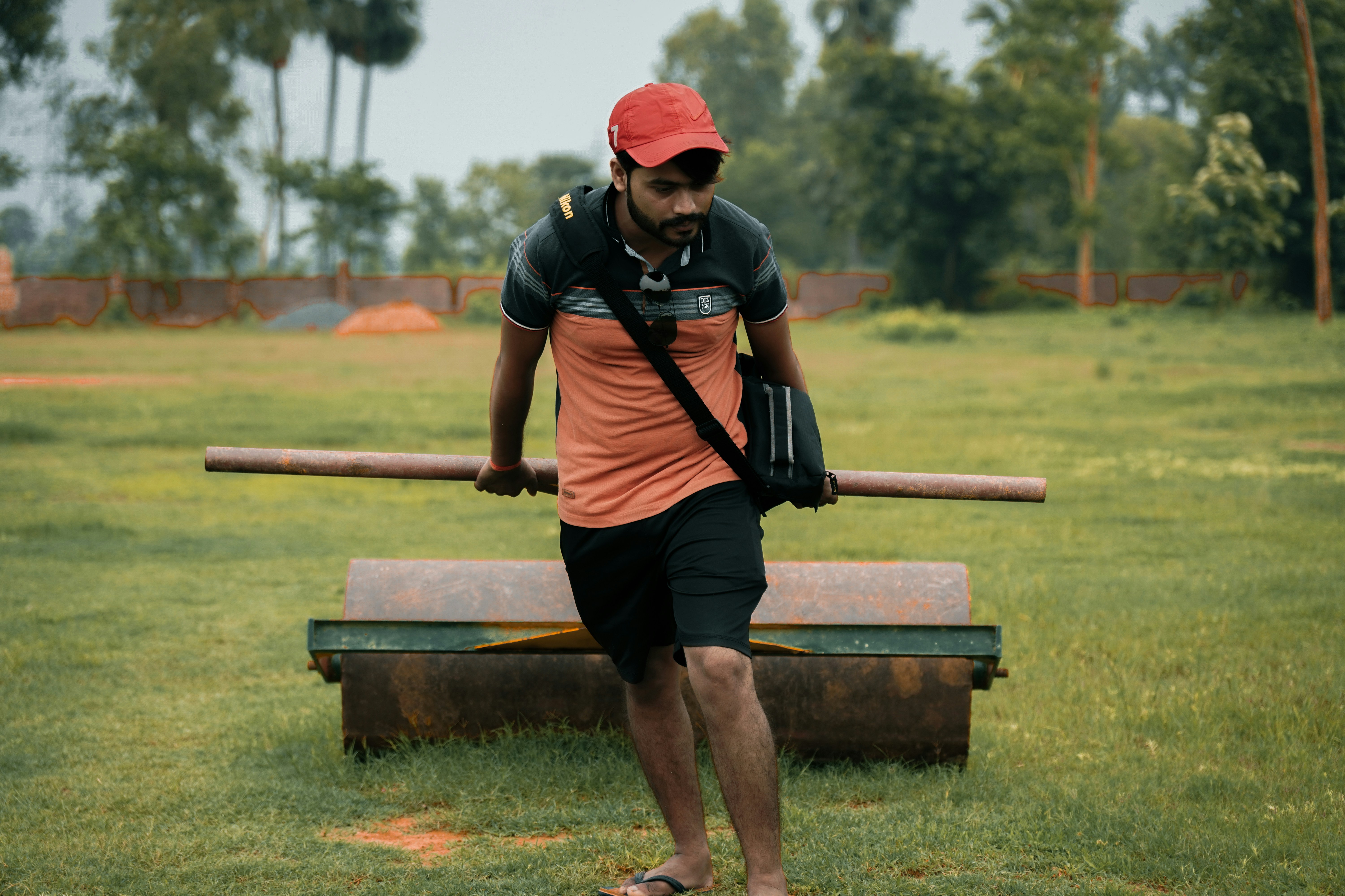 A man in a red cap carries a wooden pole across a green field, showcasing the labor of outdoor work. A roller sits in the background, hinting at agricultural activity.