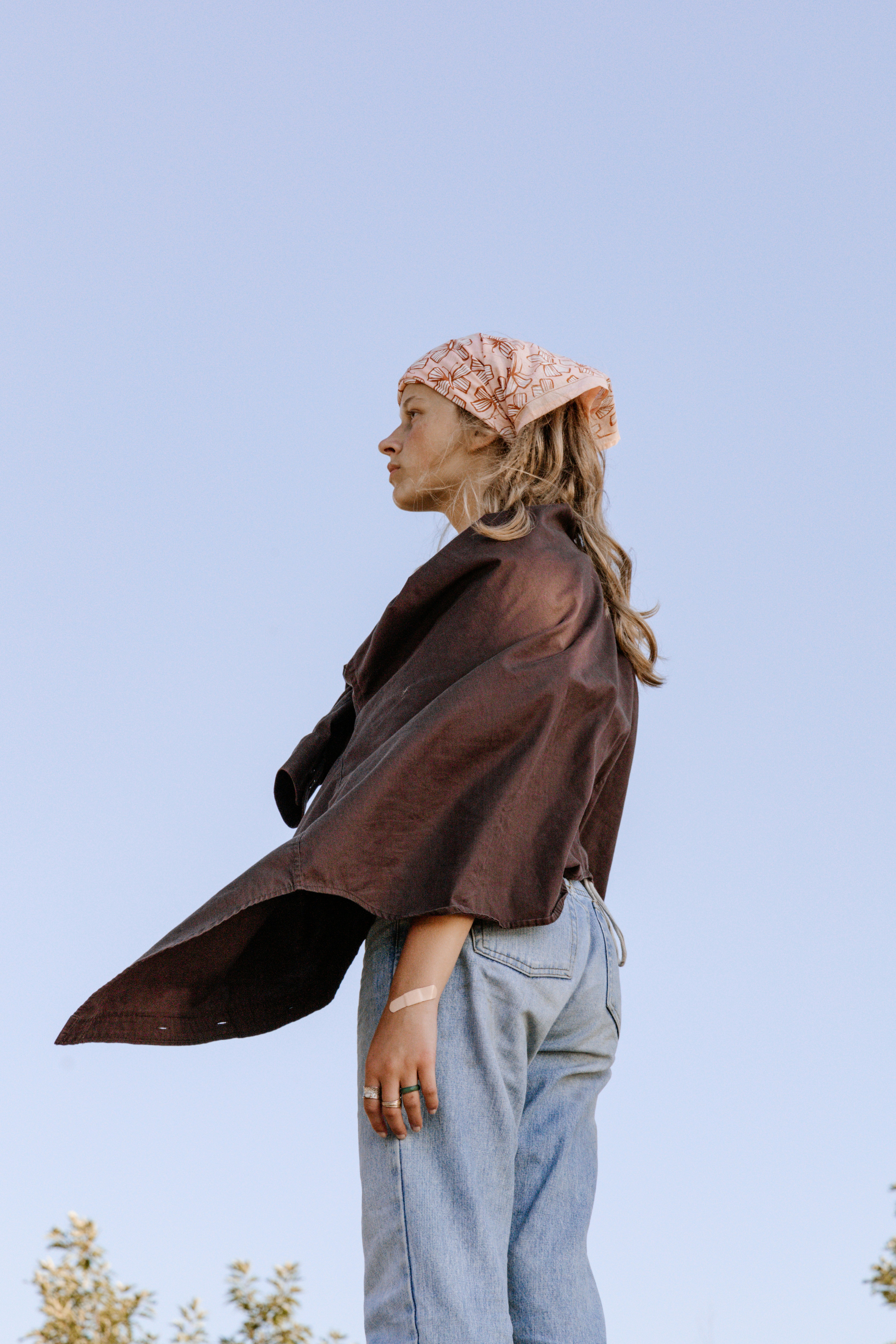 A young woman in a brown cape and pink bandana stands against a clear blue sky, exuding a sense of freedom and style.