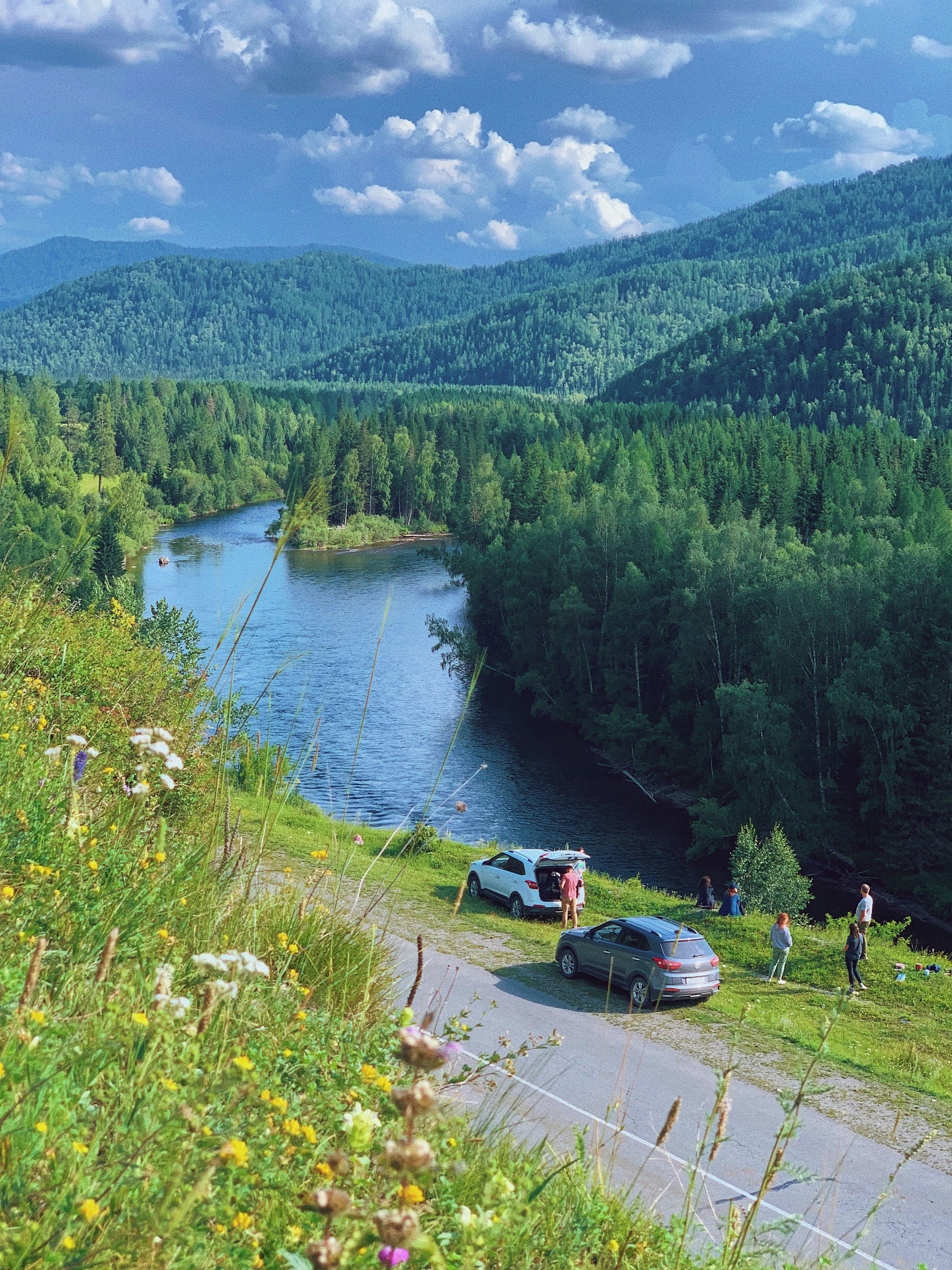 Lush green hills surround a winding river, with people enjoying leisure activities along the banks. A few cars are parked nearby, adding a sense of adventure to the tranquil scene.