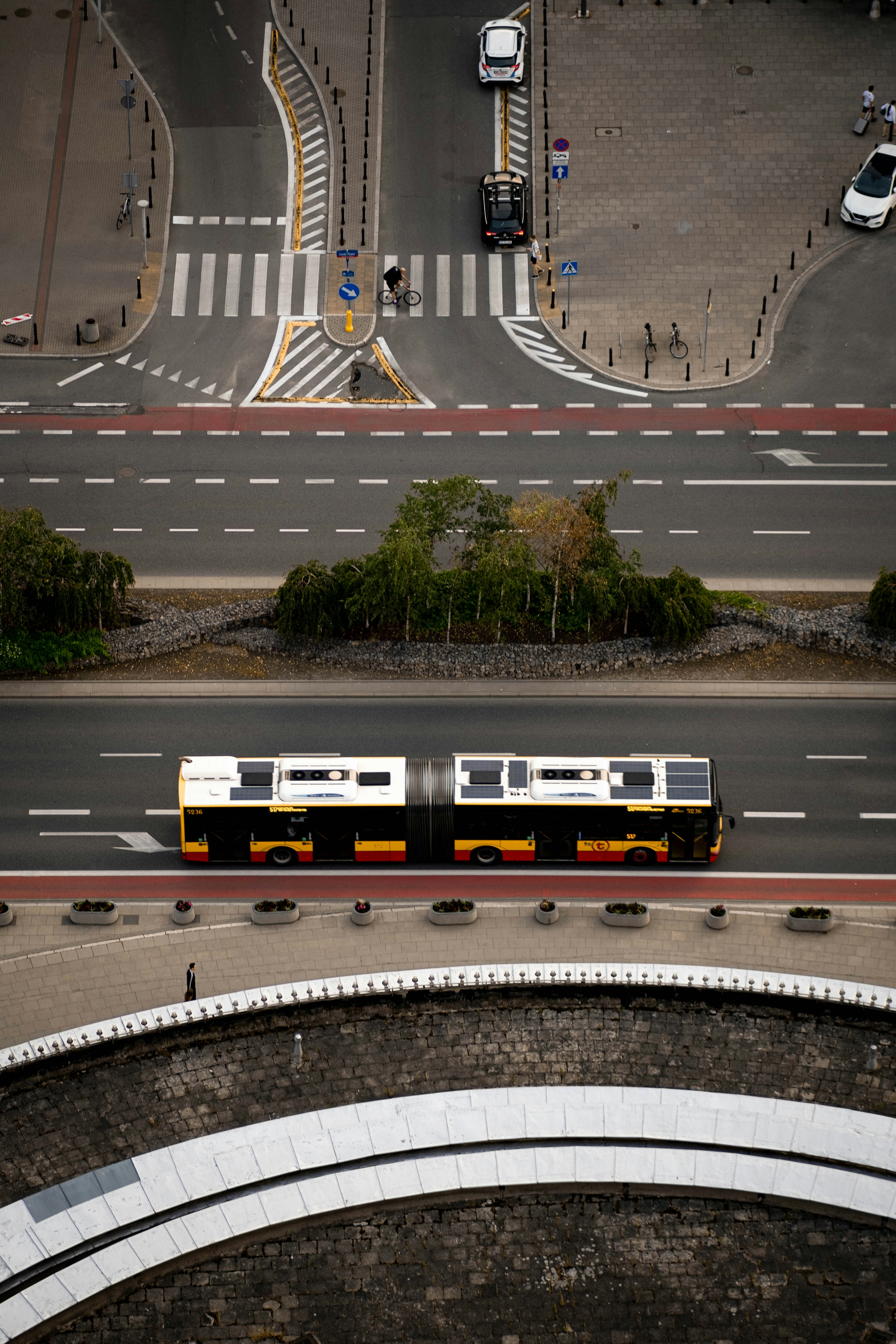 Aerial view of a bus navigating a busy urban intersection, surrounded by pedestrians and vehicles. The unique circular architecture frames the scene.
