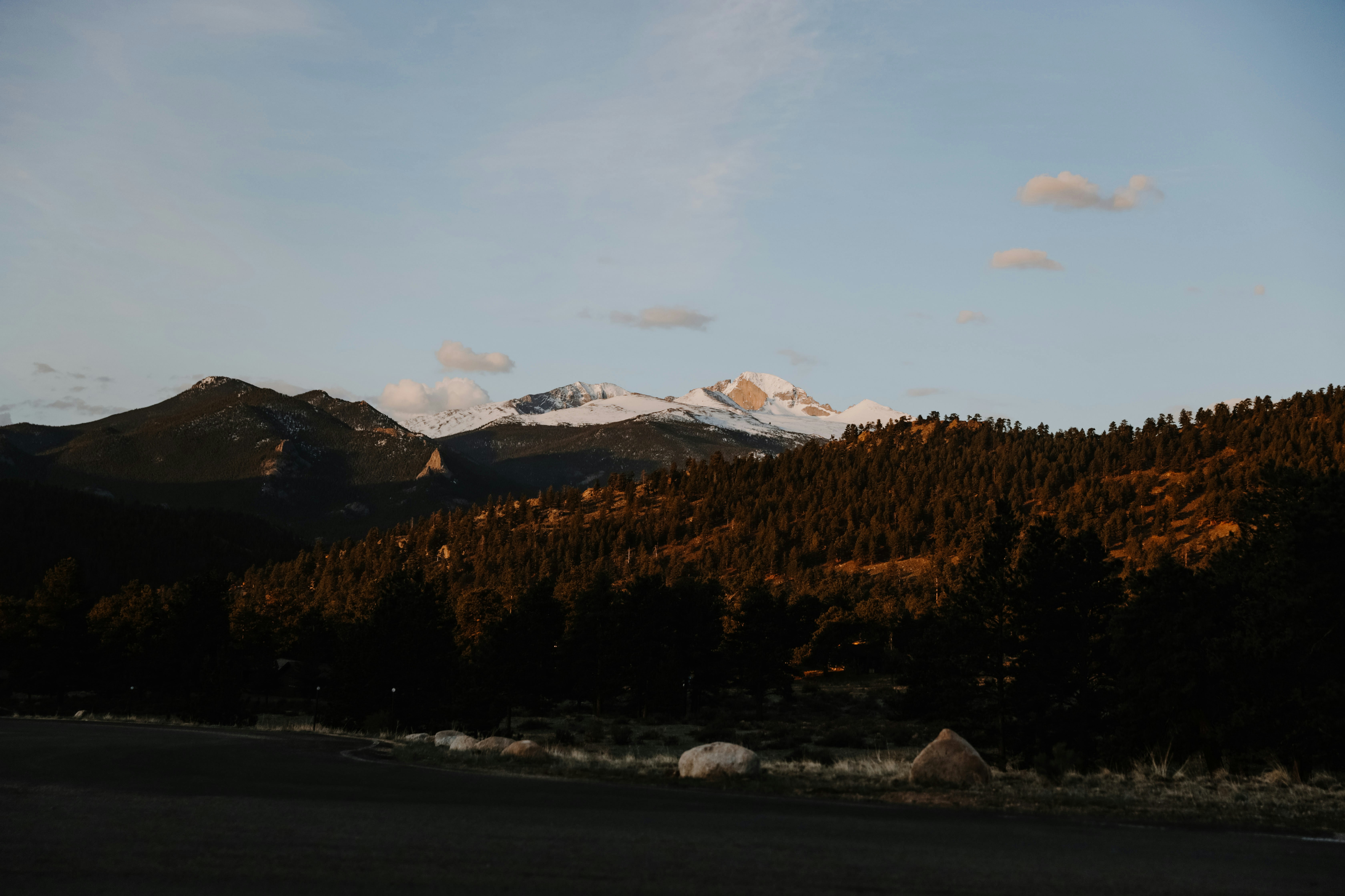 Golden light bathes the rugged mountains, highlighting the snow-capped summits against a backdrop of darkening pines. A serene landscape captures the transition from day to night.