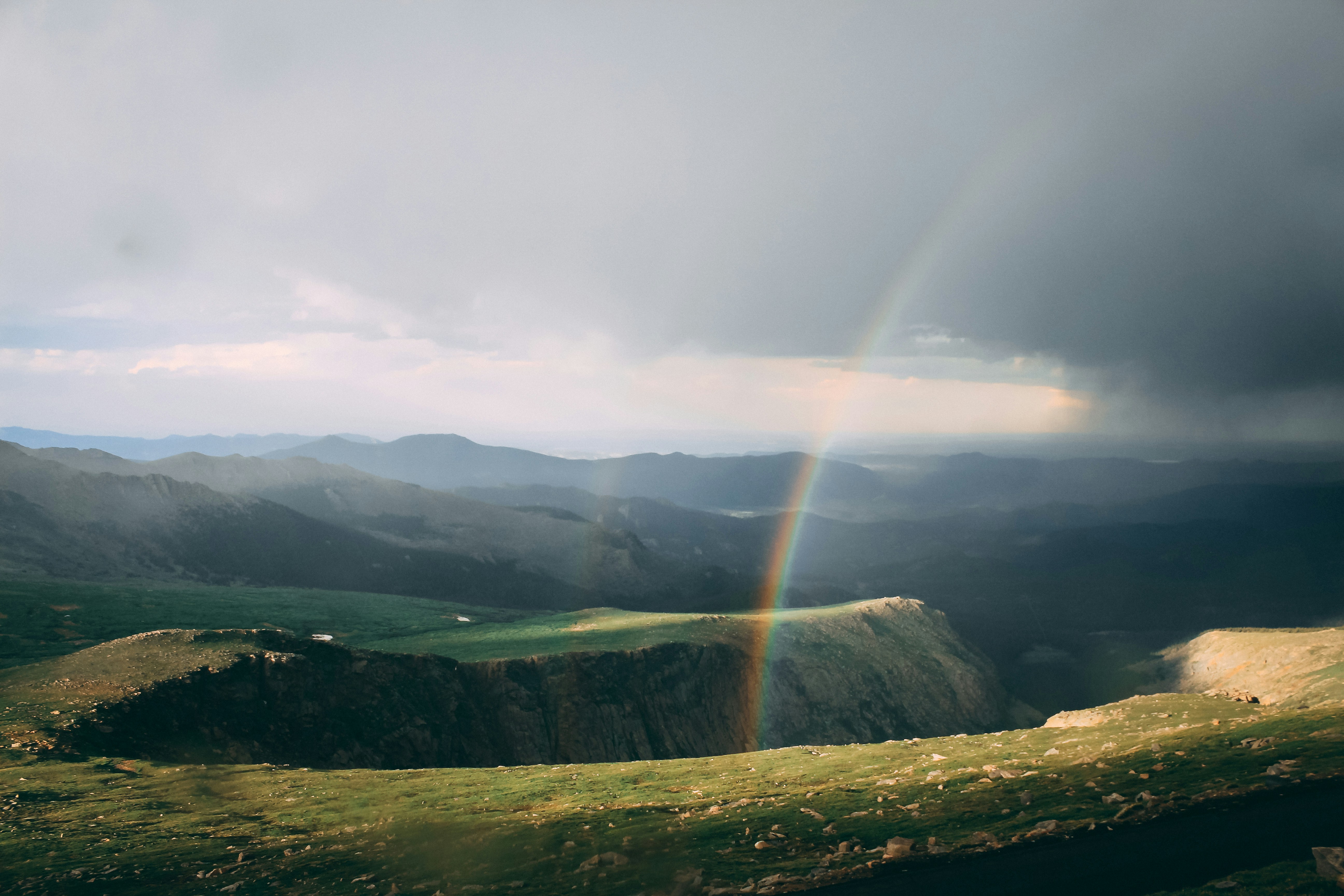A vibrant rainbow arcs over a rugged mountain landscape, contrasting with dark storm clouds in the sky. The scene captures the interplay of light and shadow across the terrain.