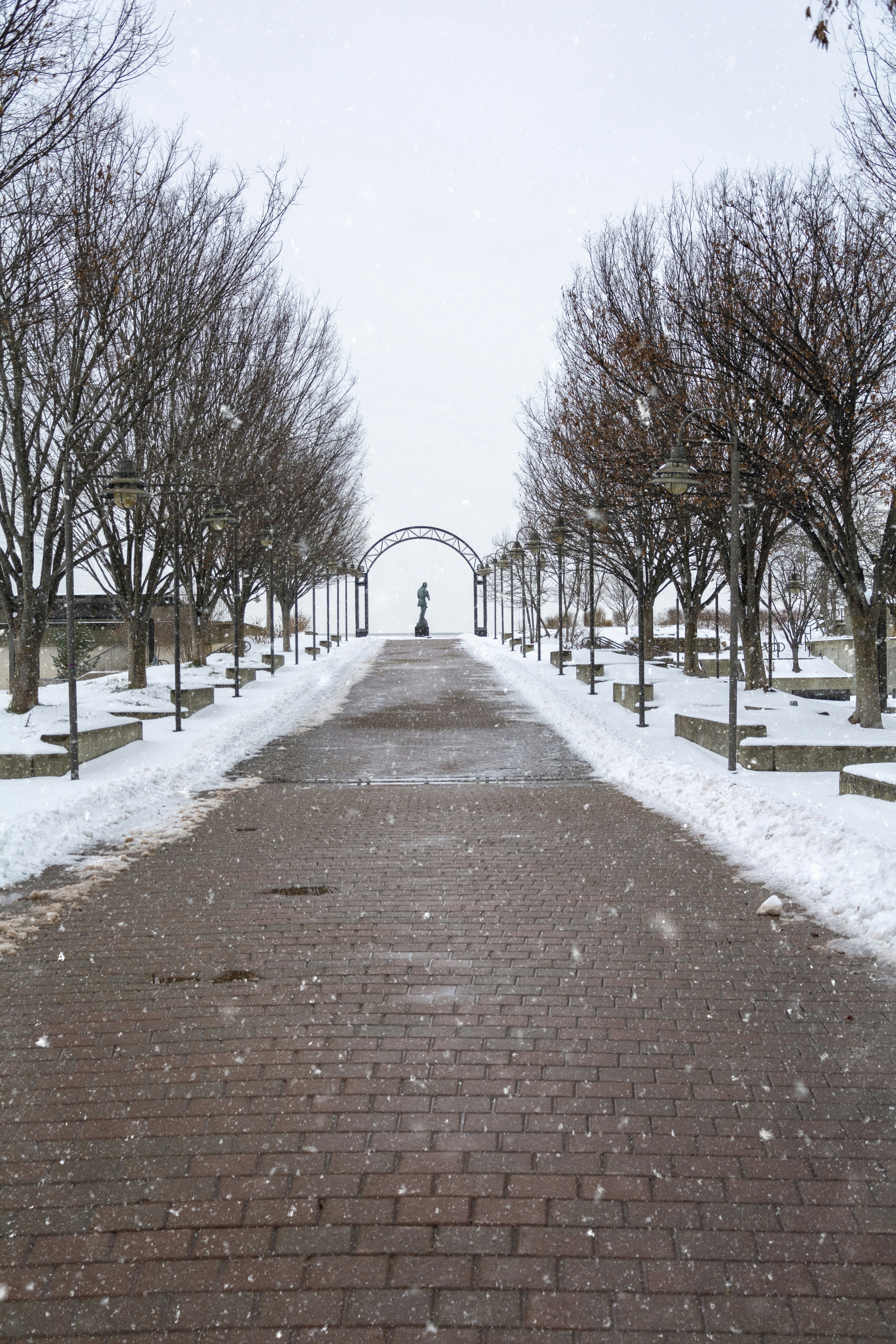 Snowbound Promenade: Brick Path Toward a Quiet Arch and Statue
