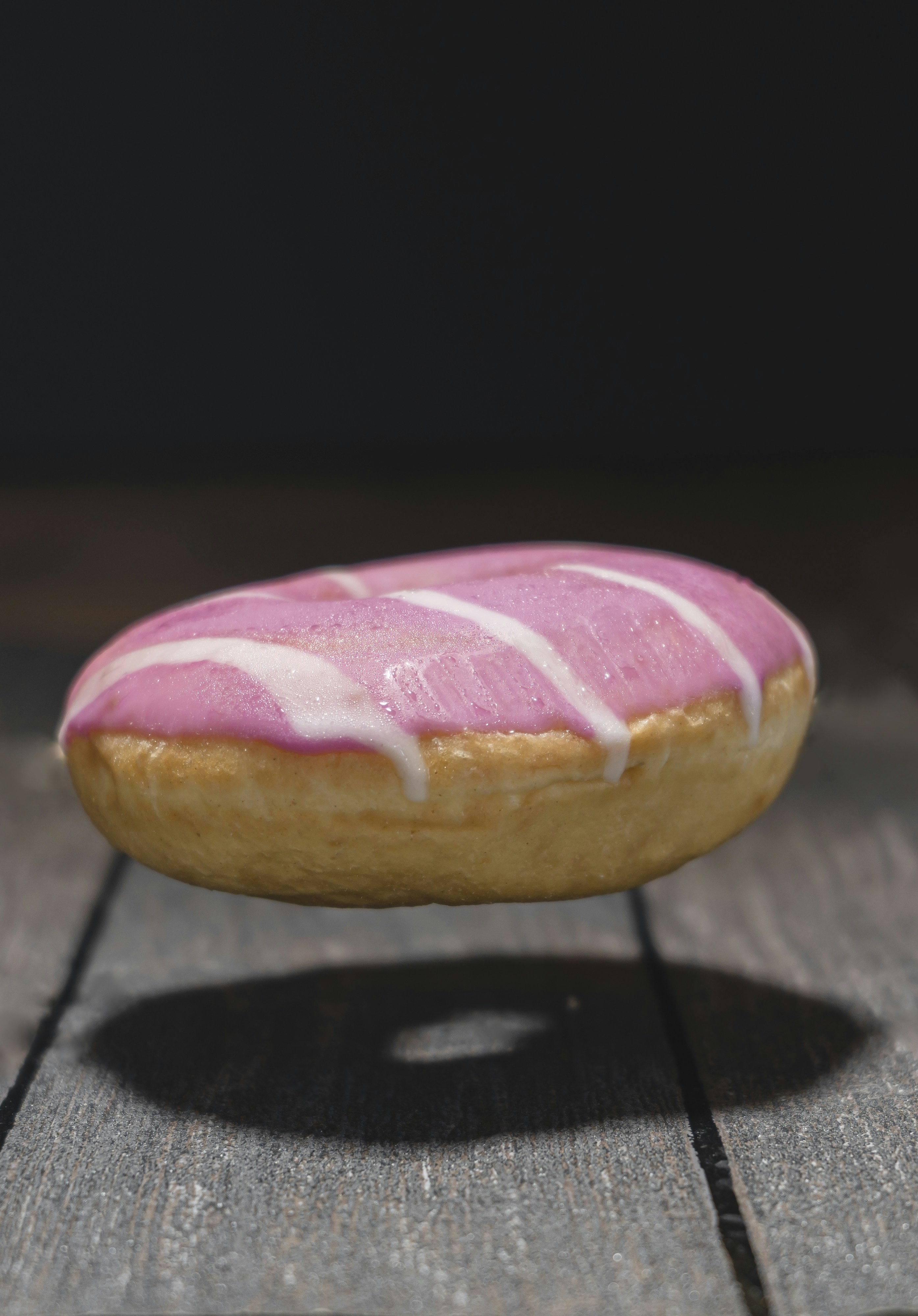A pink frosted donut suspended mid-air, casting a soft shadow on a rustic wooden surface.