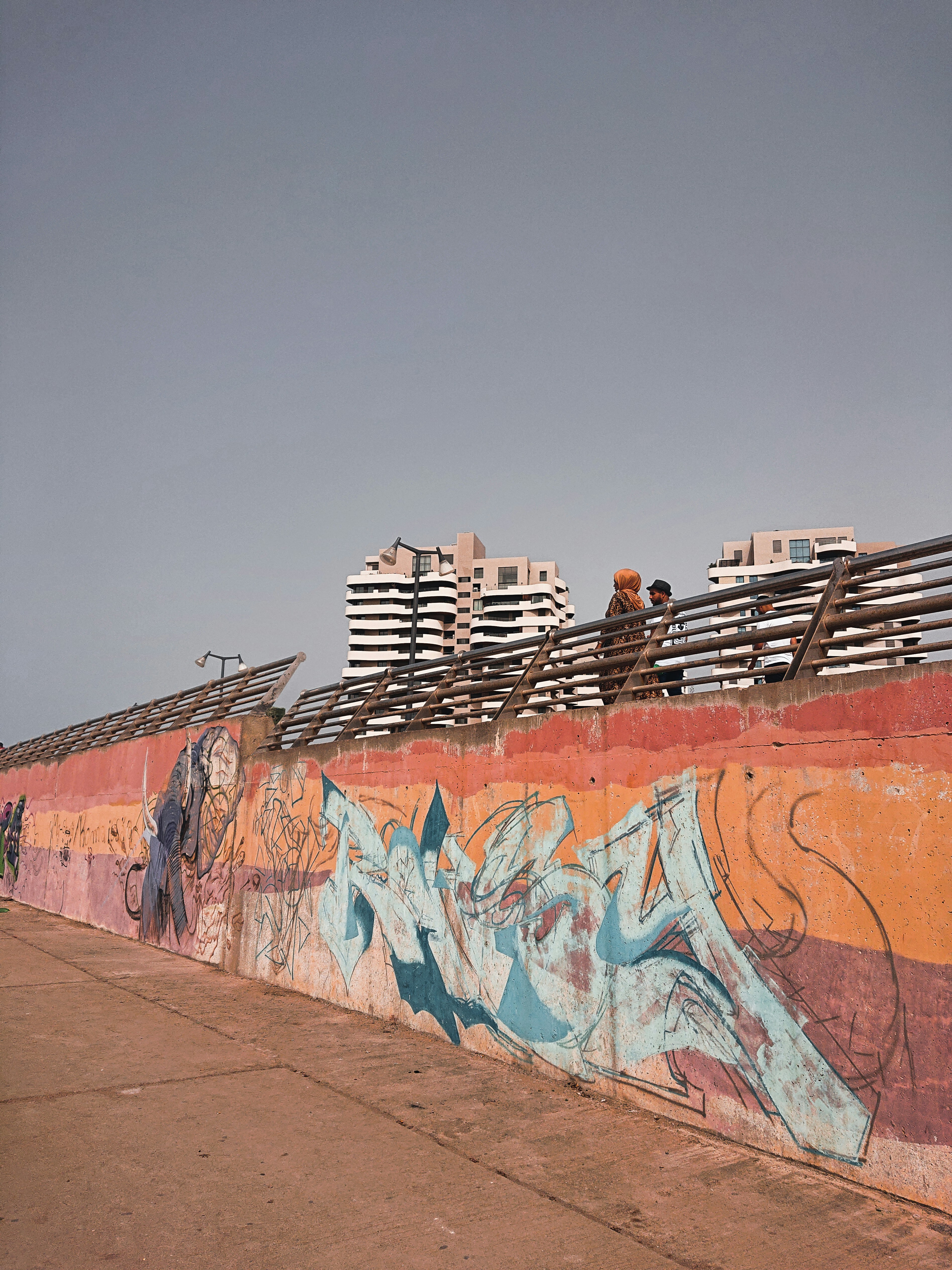 Vibrant graffiti adorns a concrete wall under a modern urban skyline, where two figures engage in conversation. The scene captures the intersection of street art and contemporary architecture.