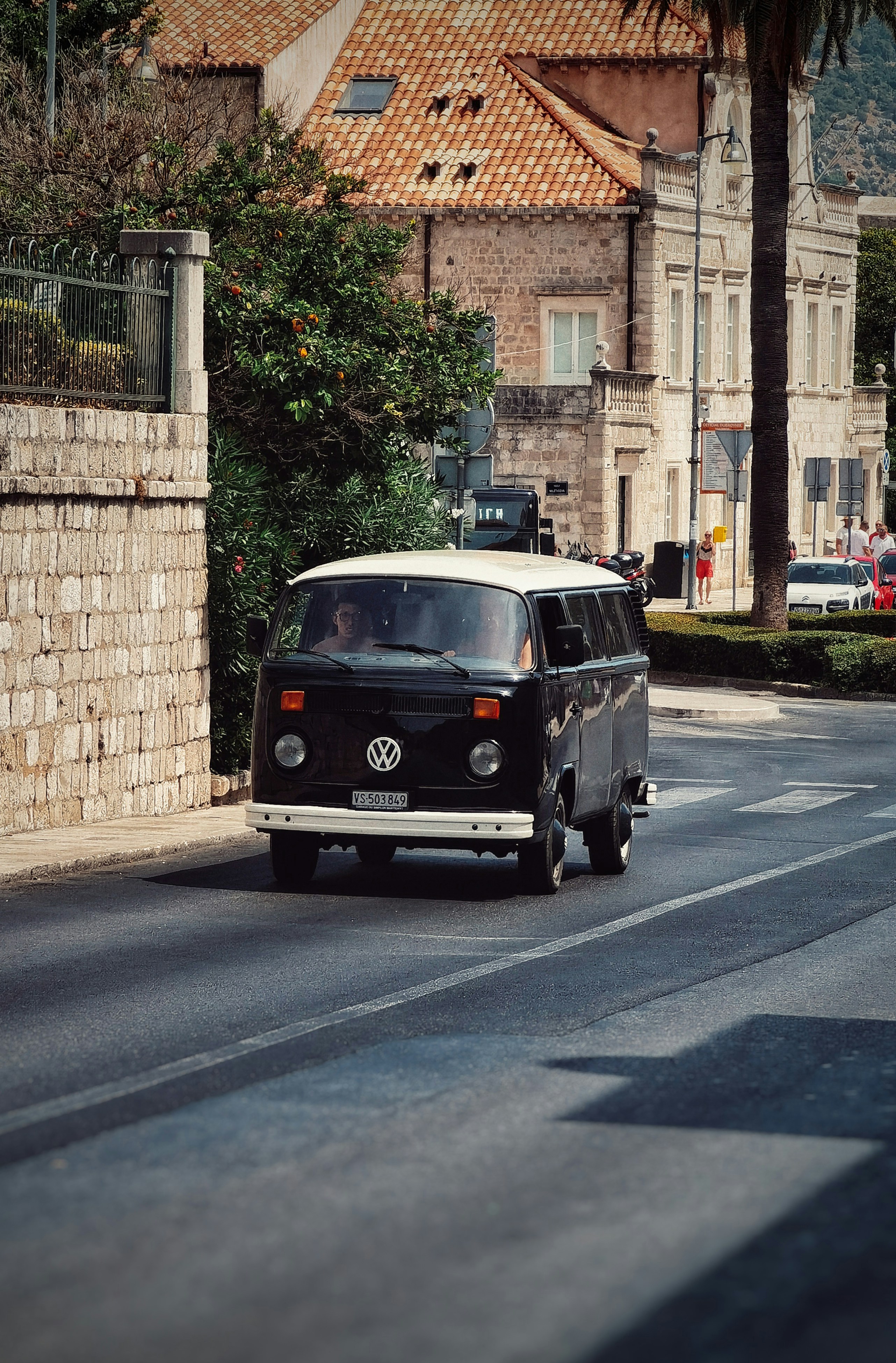 A vintage Volkswagen van cruising along a picturesque street, surrounded by historic architecture and lush greenery.