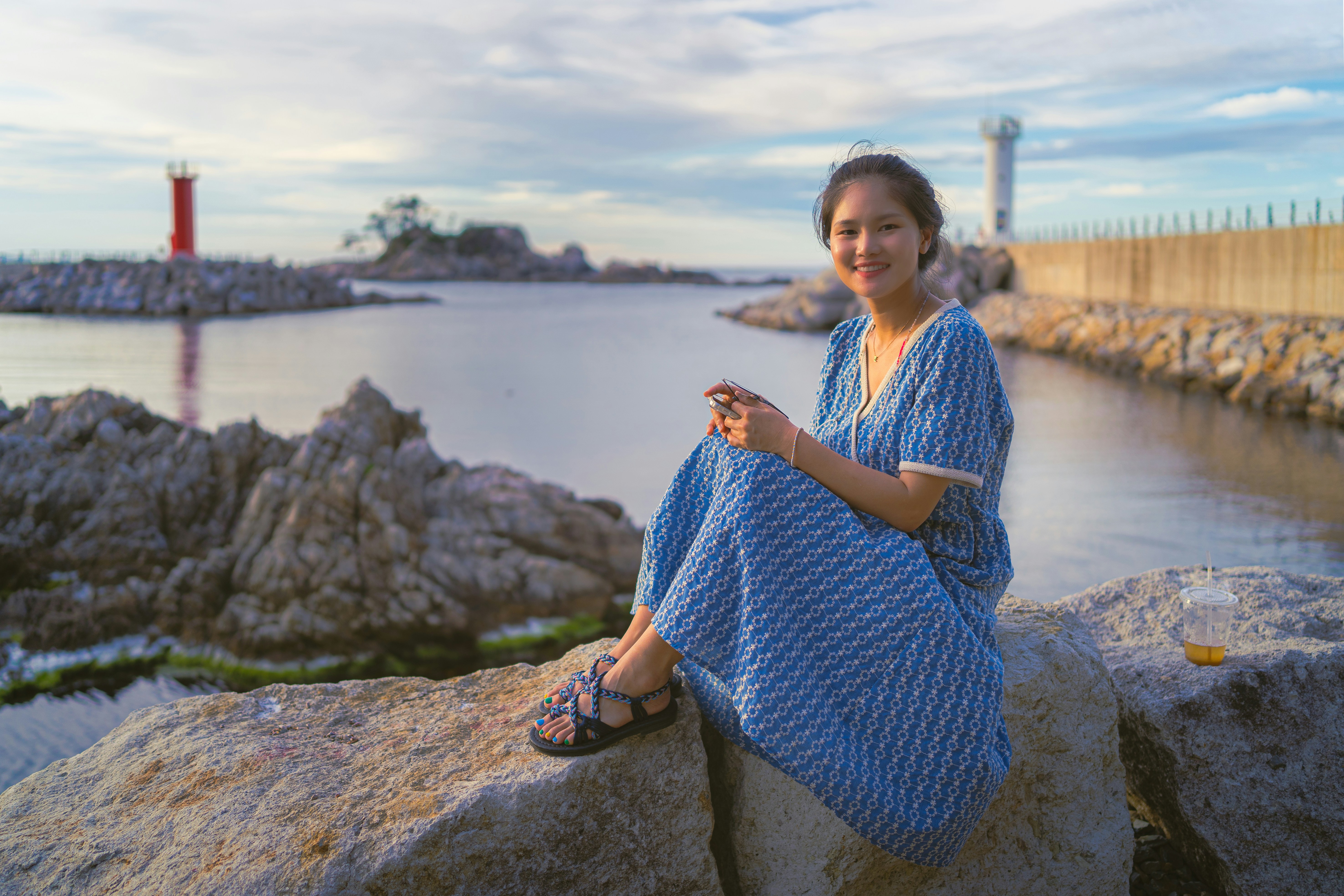 A girl in a blue dress sitting in front of a harbor with a lighthouse.
